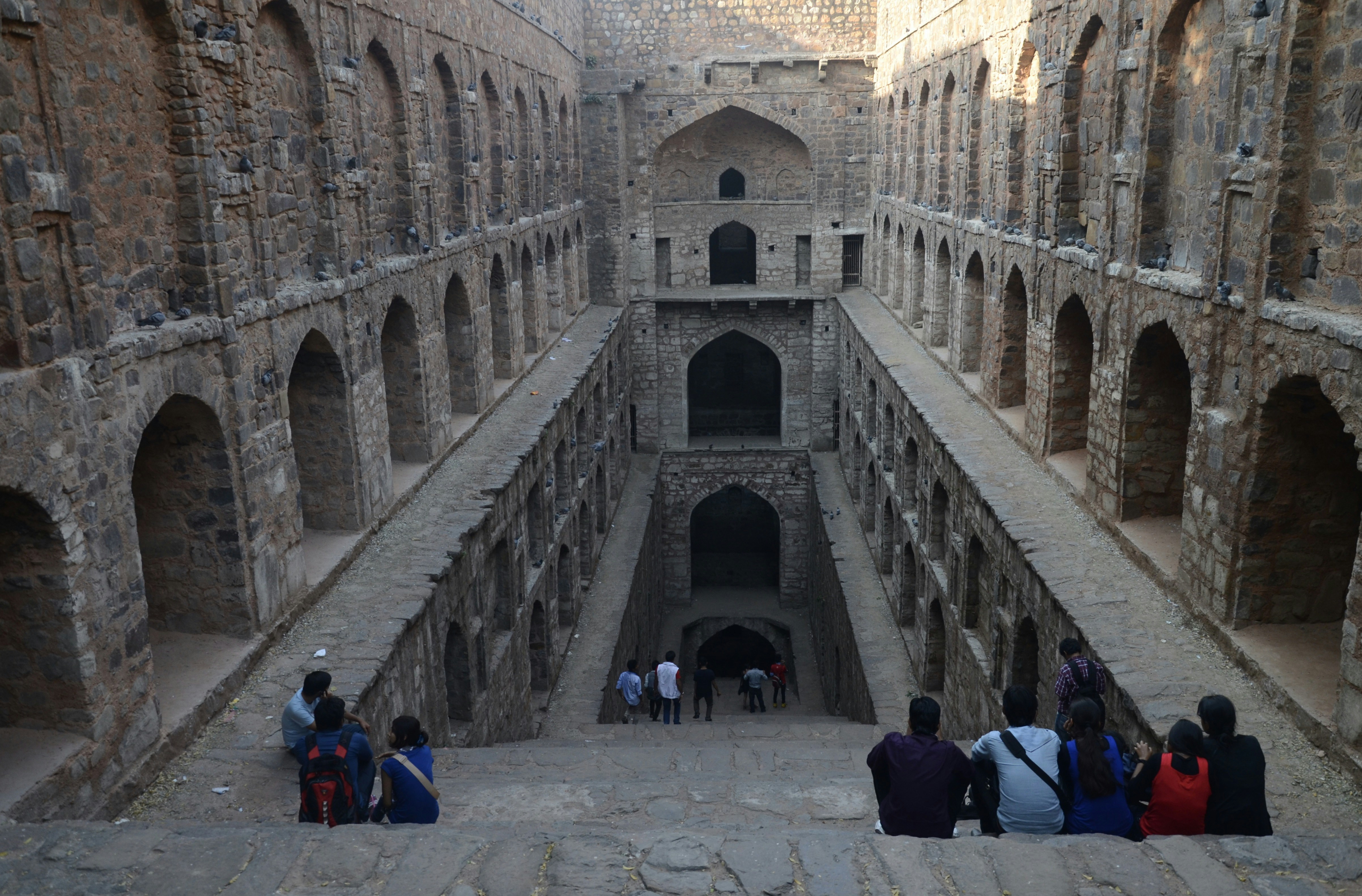Ancient stone stepwell with descending symmetrical arches and people sitting on its edges.