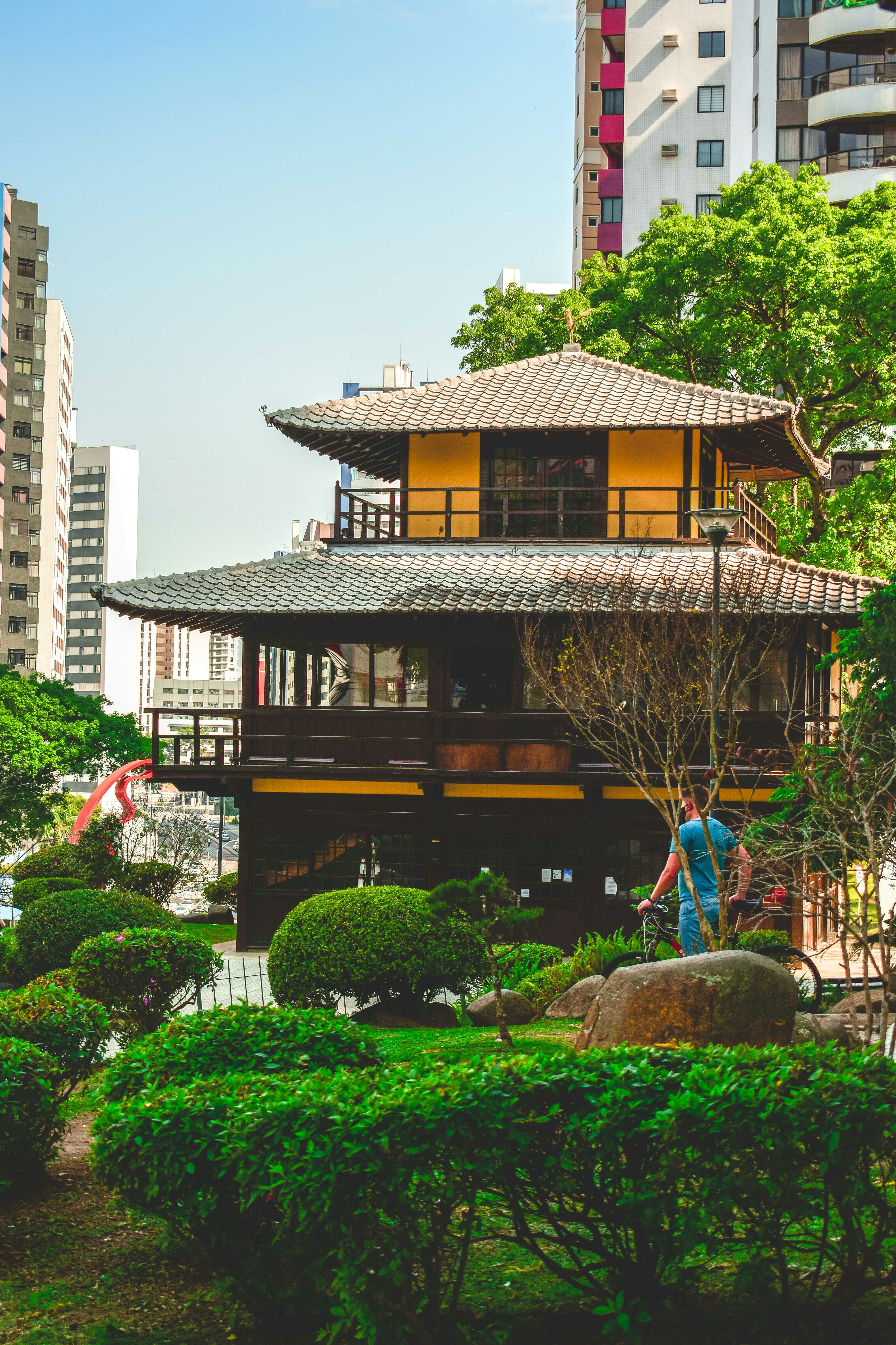 Traditional Japanese-style house nestled in a lush garden, surrounded by modern skyscrapers. A person interacts with the landscape, highlighting the blend of nature and architecture.