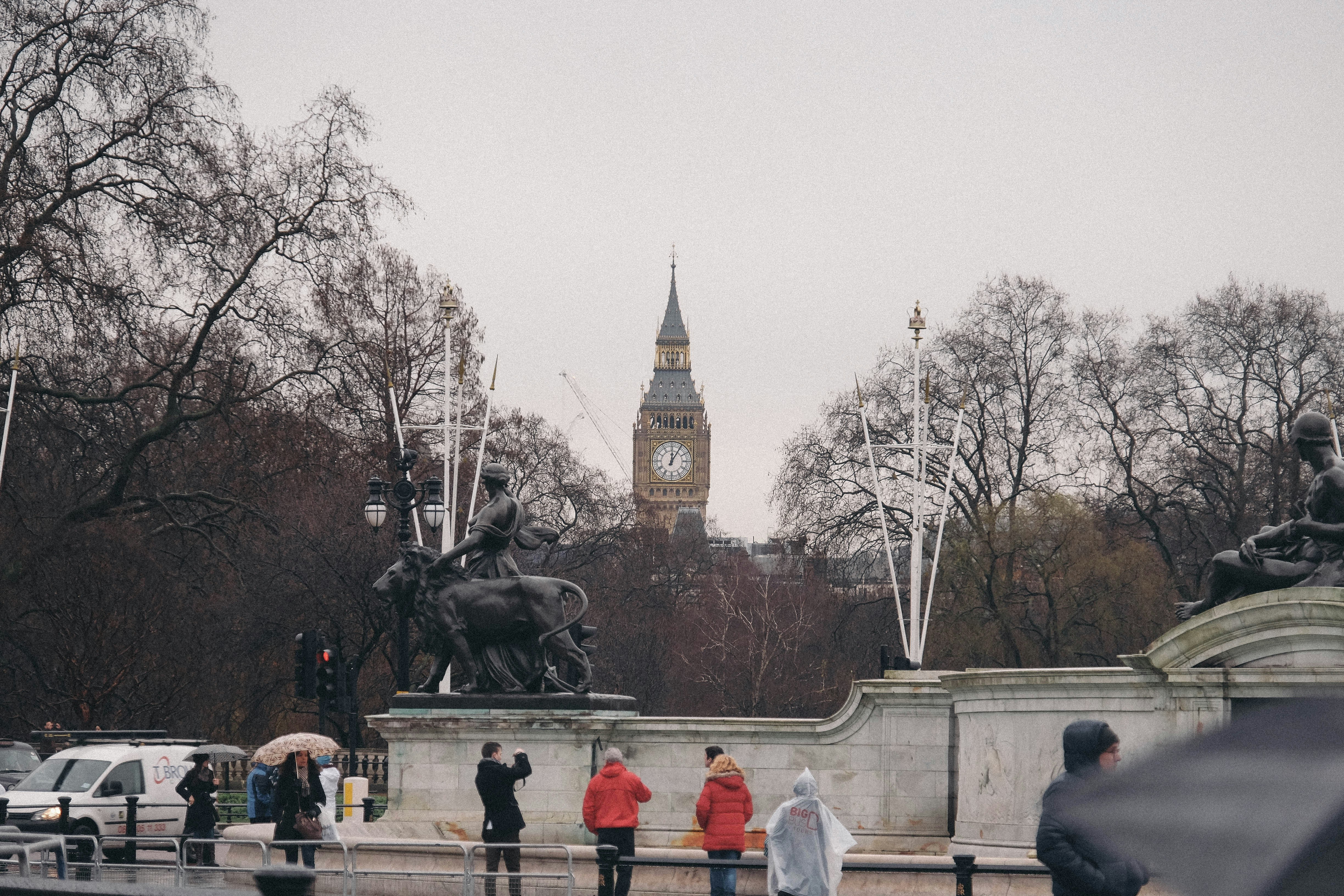 Iconic clock tower stands tall amidst a bustling scene, framed by statues and trees in a gray, overcast setting.