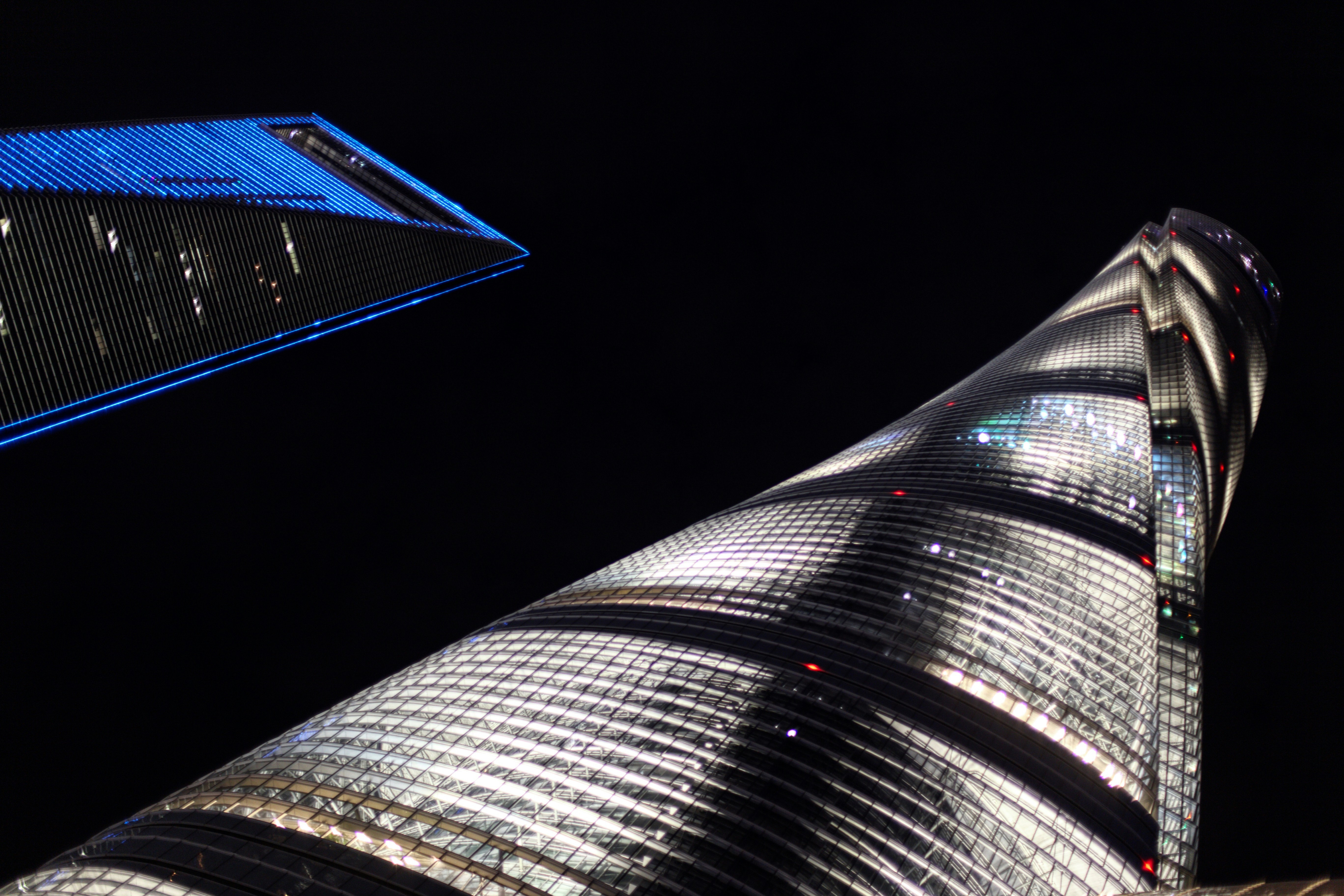 Two towering skyscrapers illuminated against the night sky, showcasing intricate designs and vibrant lighting. The perspective emphasizes their grandeur and architectural details.