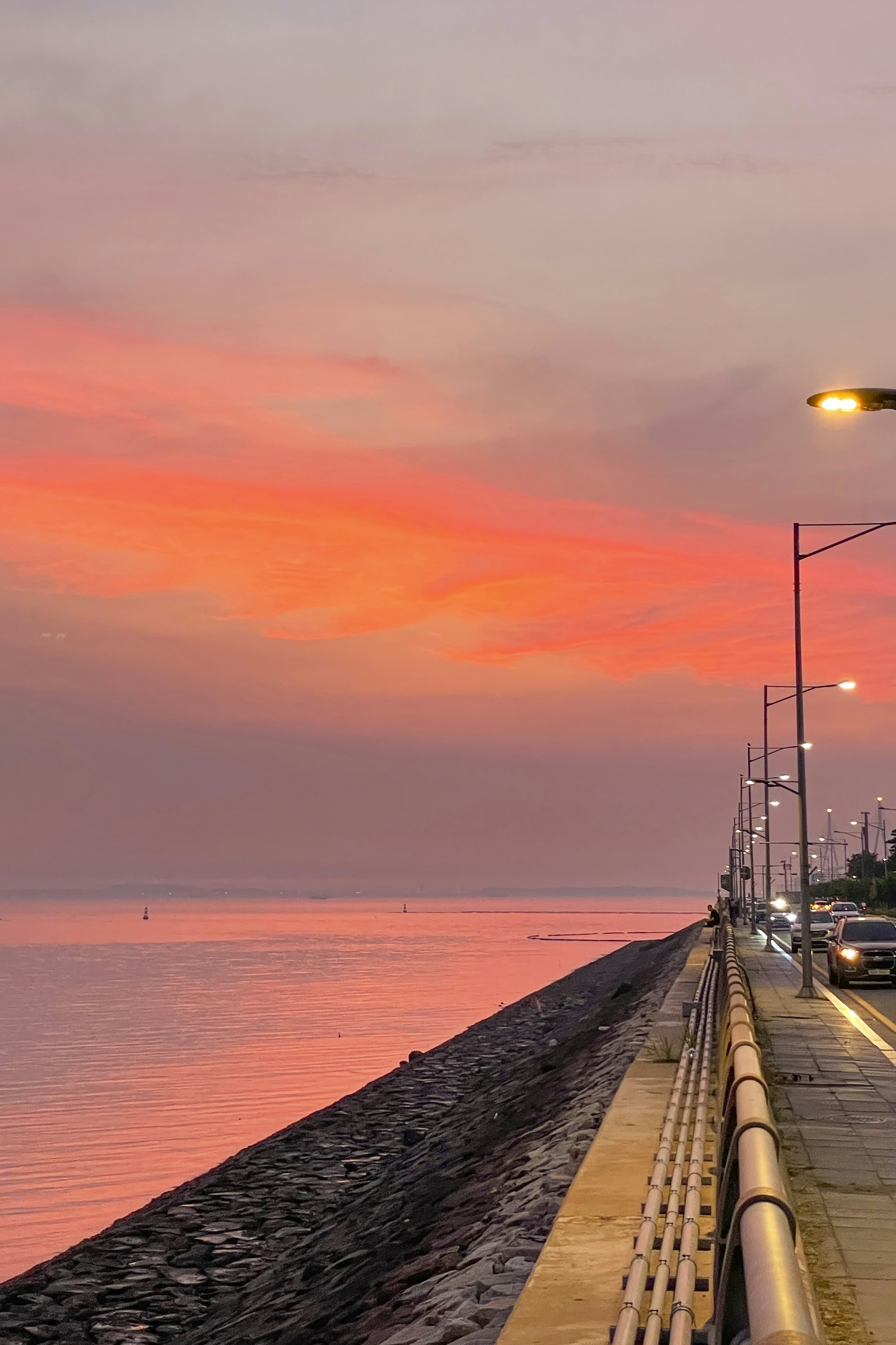 A tranquil waterfront scene at sunset, showcasing a path lined with streetlights and a calm sea reflecting vibrant hues of orange and pink.