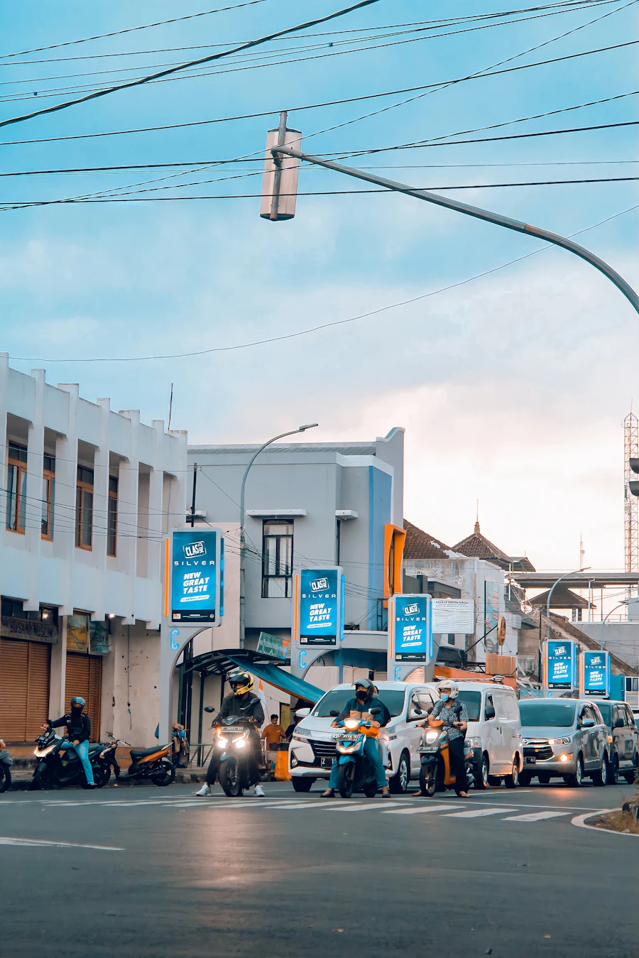 Motorcycles and cars wait at a busy urban intersection lined with low-rise buildings and overhead power lines, under a pale blue evening sky.
