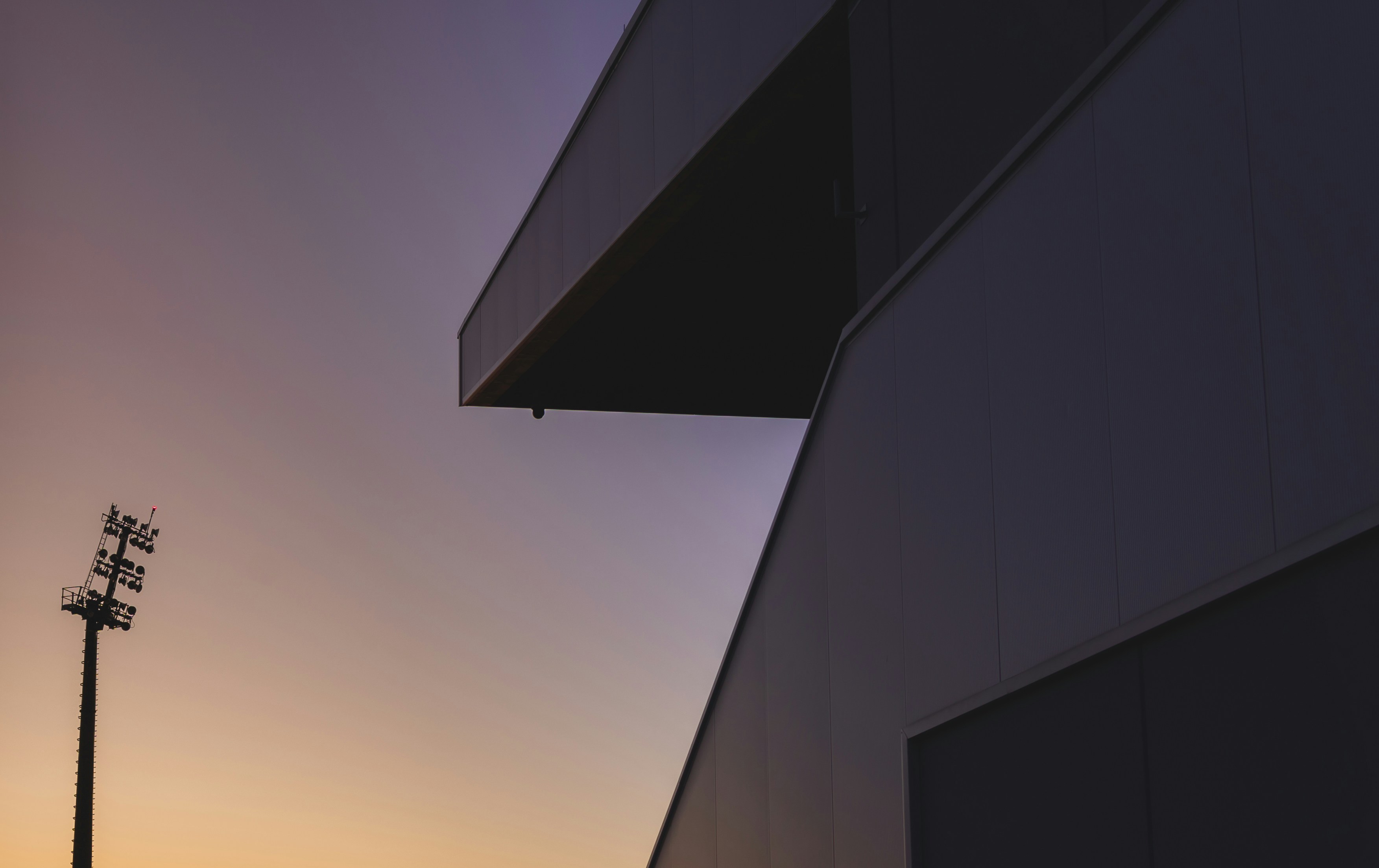 Modern building structure contrasted with a stadium floodlight at dusk, highlighting geometric lines and soft gradient sky.