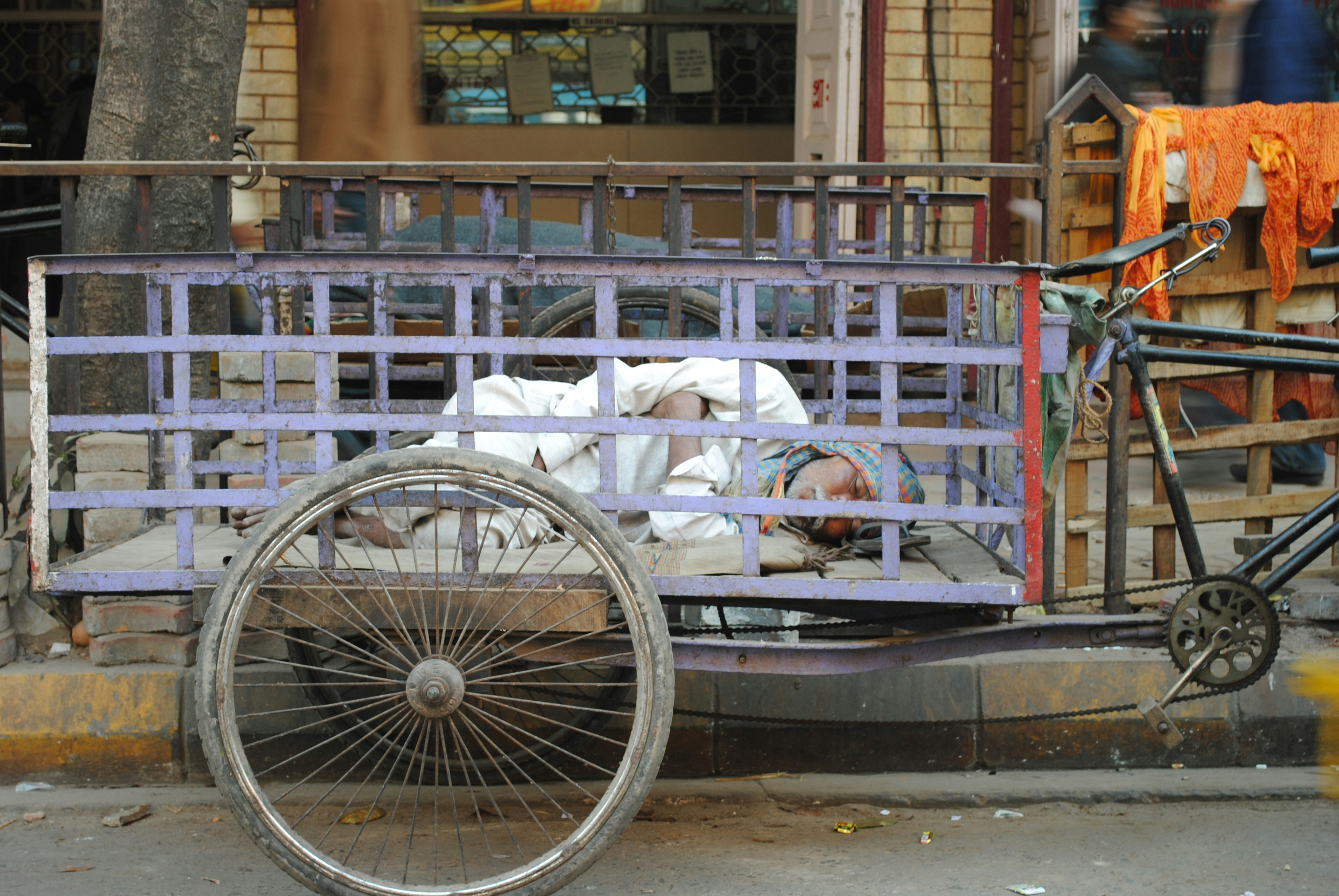 A man reclines in a colorful cart, surrounded by the vibrant hues of street life. The scene captures a moment of rest in a bustling environment.