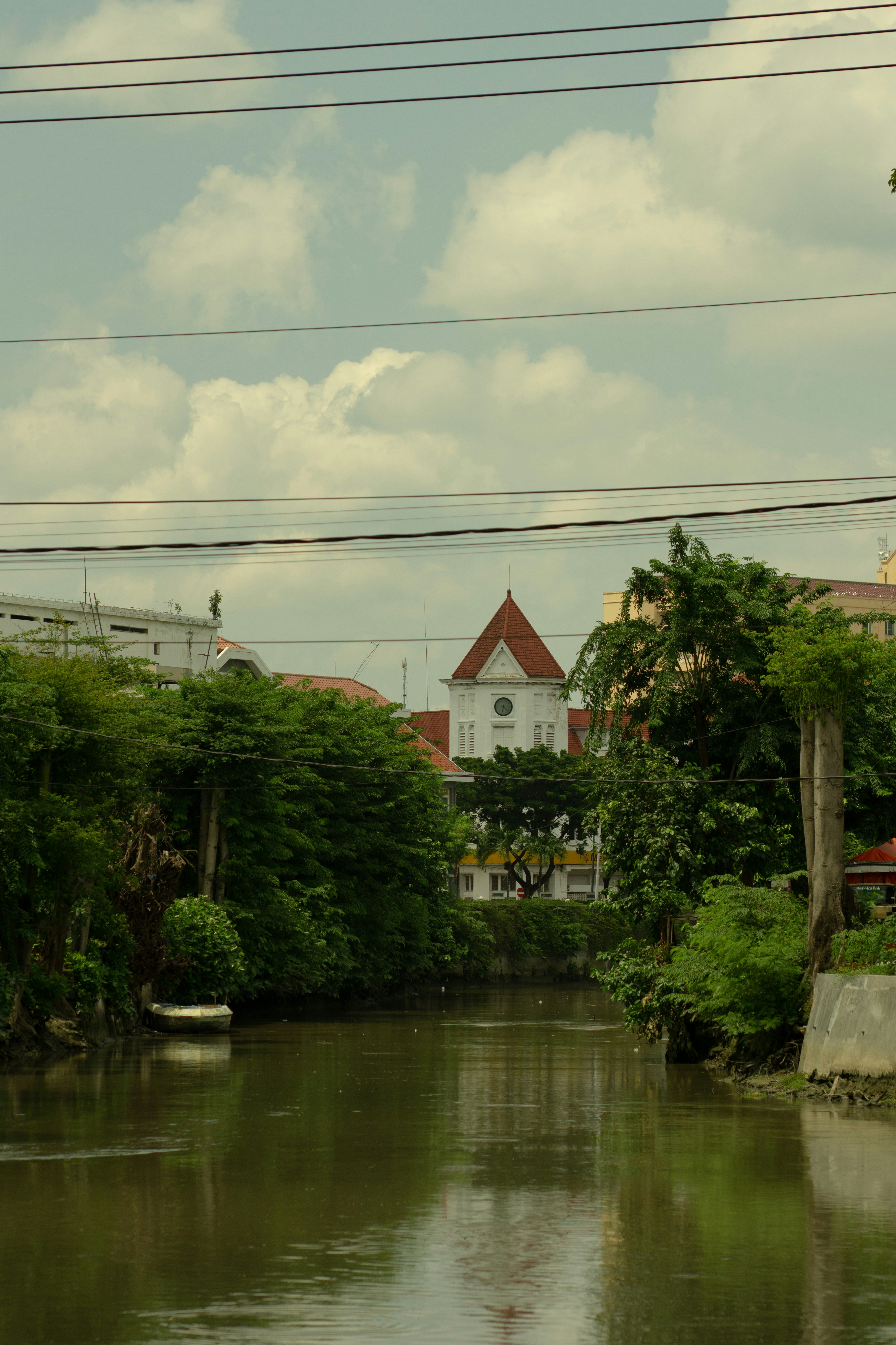 Historic building with a red-tiled roof reflected in a tranquil waterway, framed by lush greenery and power lines above.