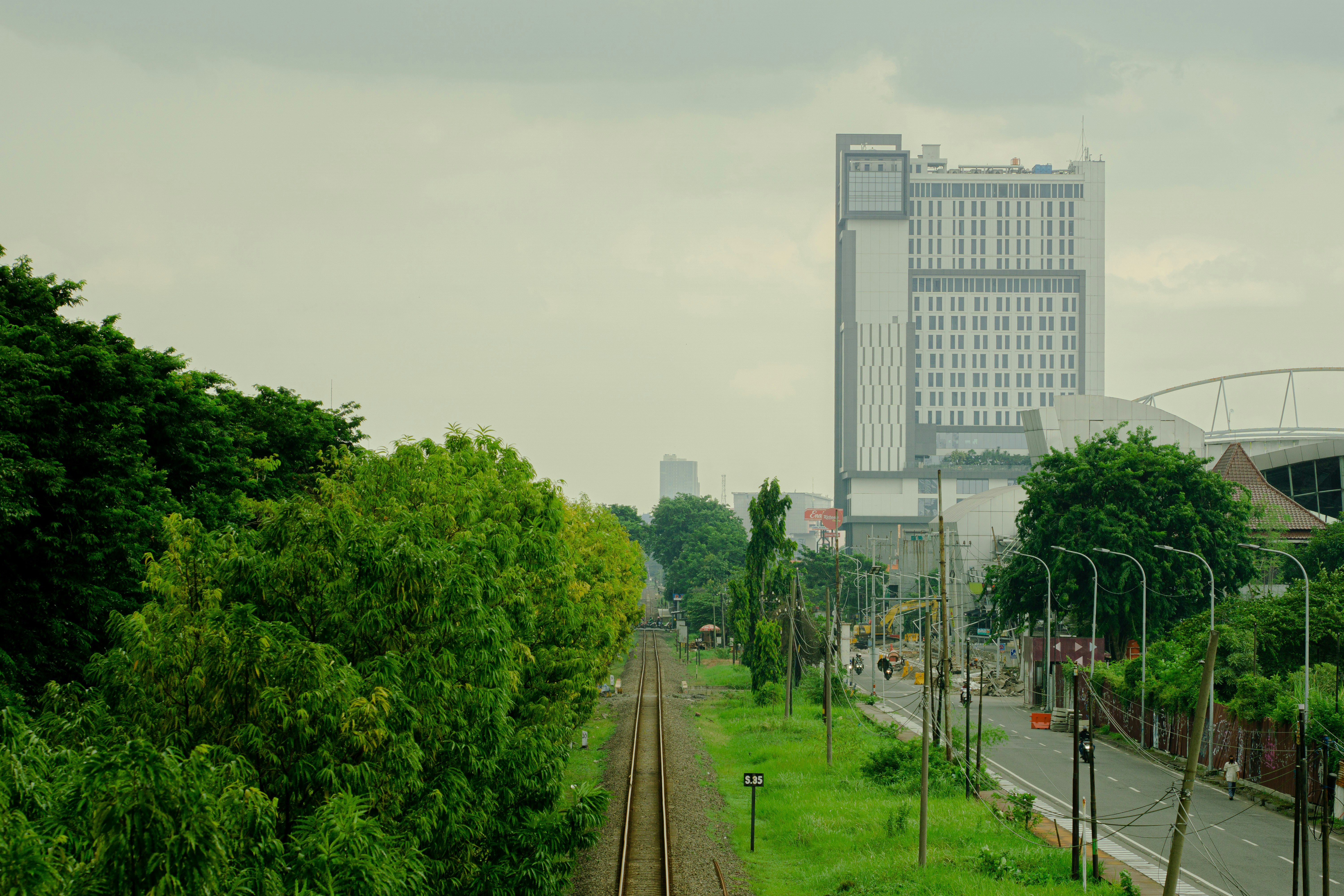 A view of railway tracks lined with lush greenery, leading towards a modern skyscraper under an overcast sky.