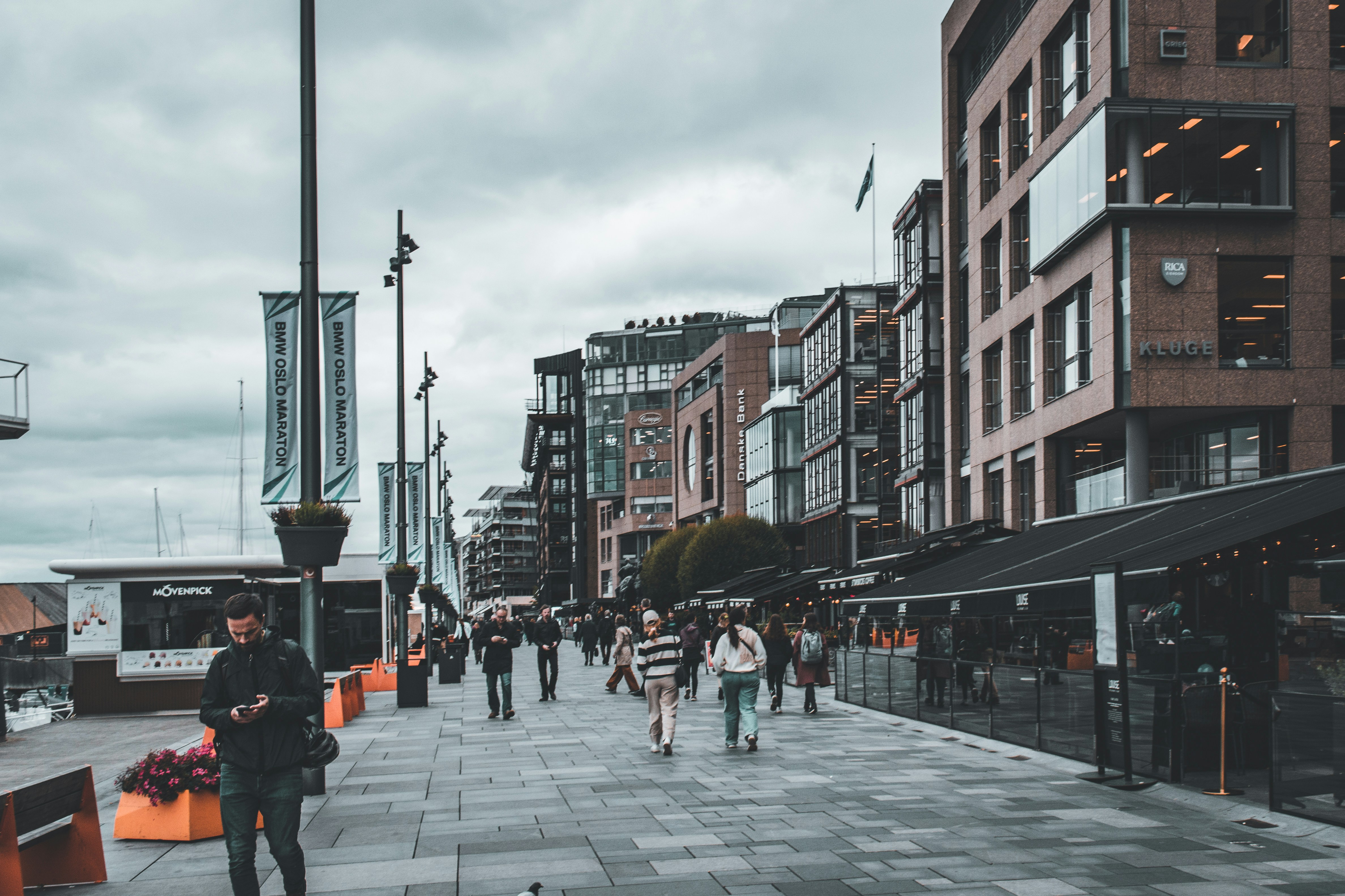 People walking along a bustling city promenade lined with modern buildings under a cloudy sky.