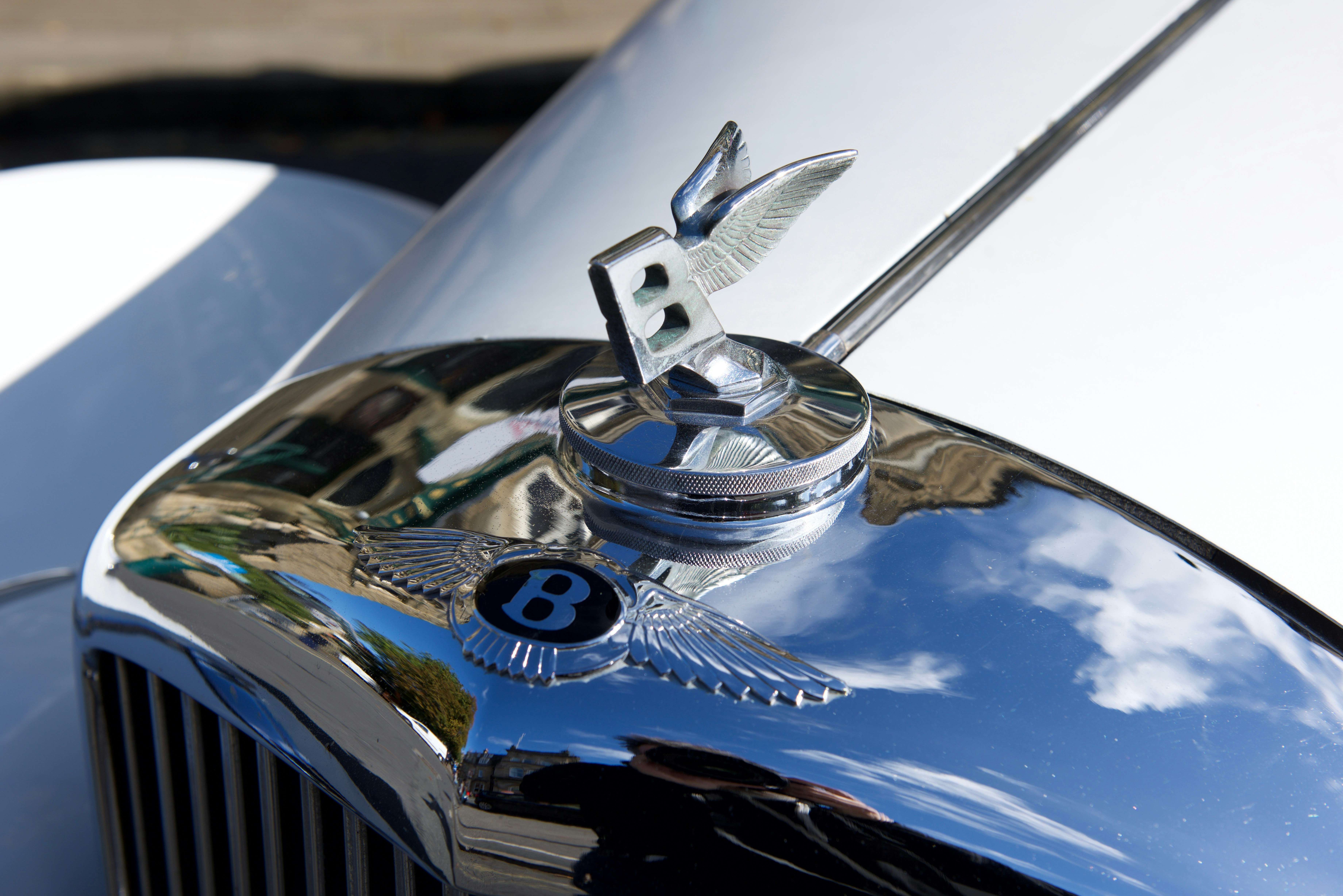 Bentley hood ornament reflecting blue sky and surrounding buildings on polished chrome surface.