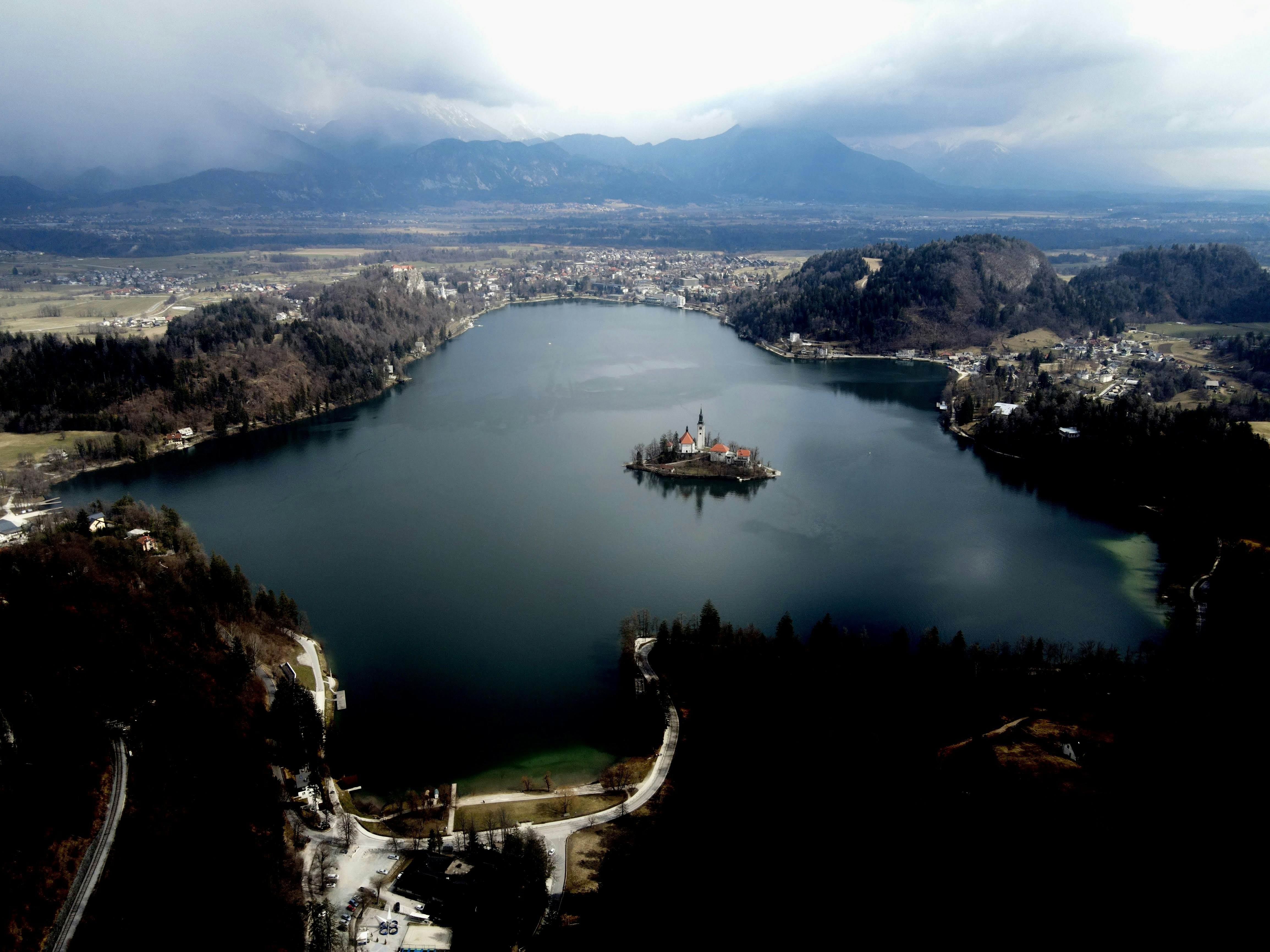 Aerial view of Lake Bled with its iconic island and church surrounded by mountainous landscape under cloudy skies.