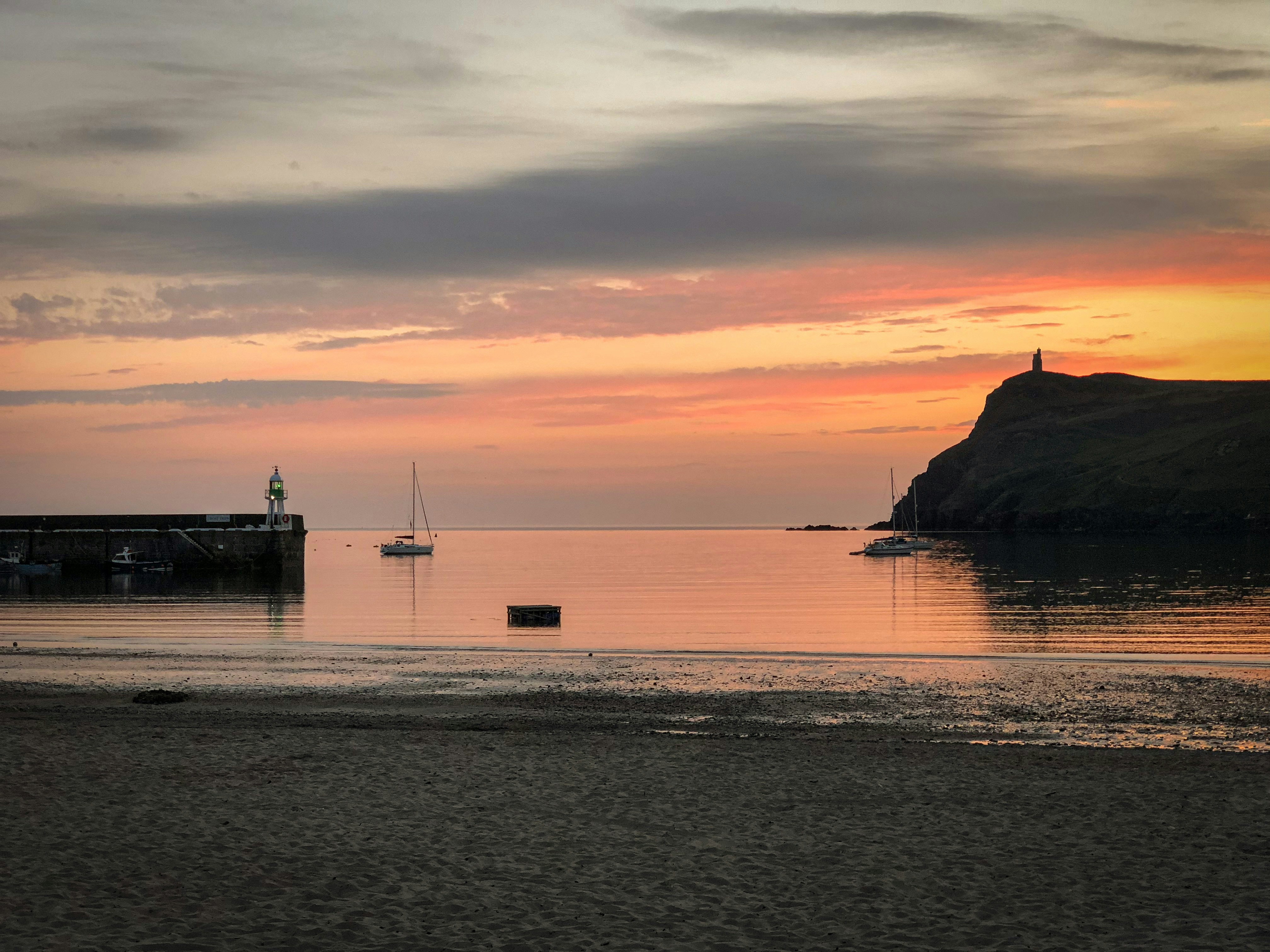 Sailboats anchored in calm waters under a vibrant sunset sky with a silhouette of a hill in the background.