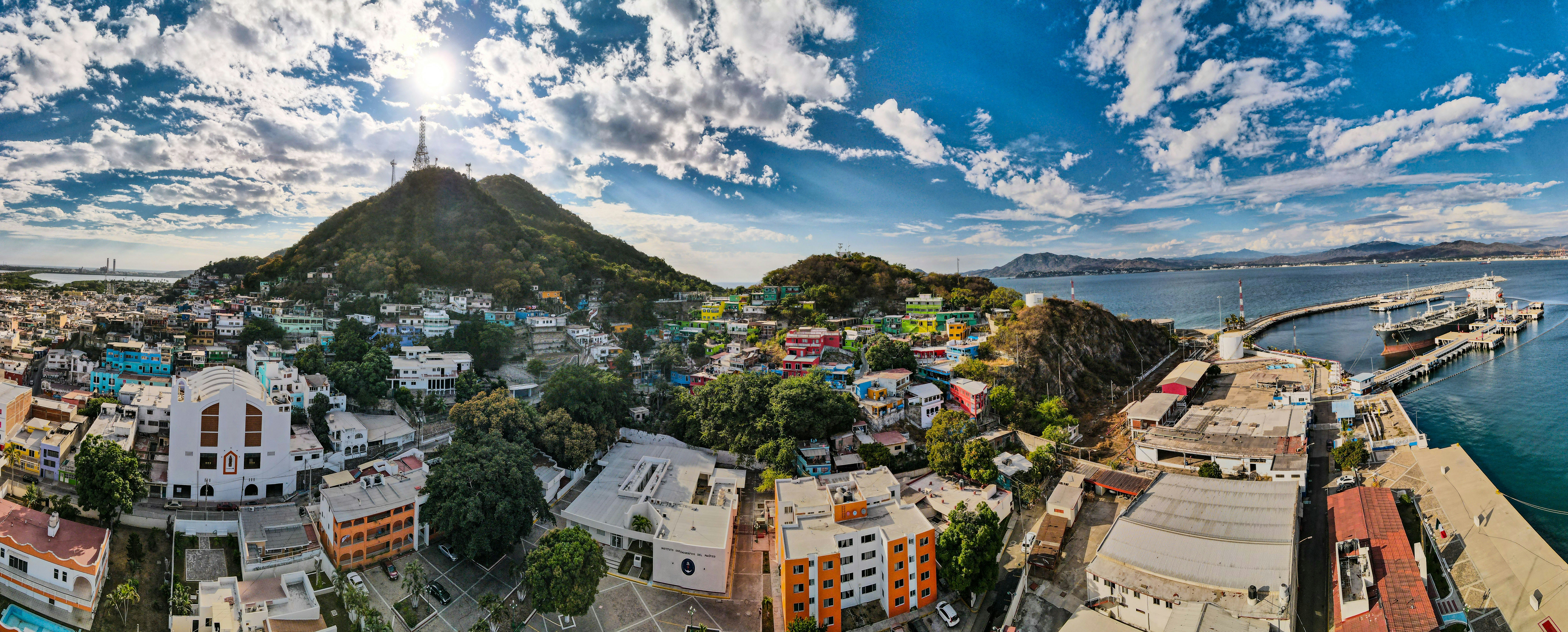 Panoramic view of a coastal town with colorful buildings, a mountainous backdrop, and a bright sky filled with clouds.