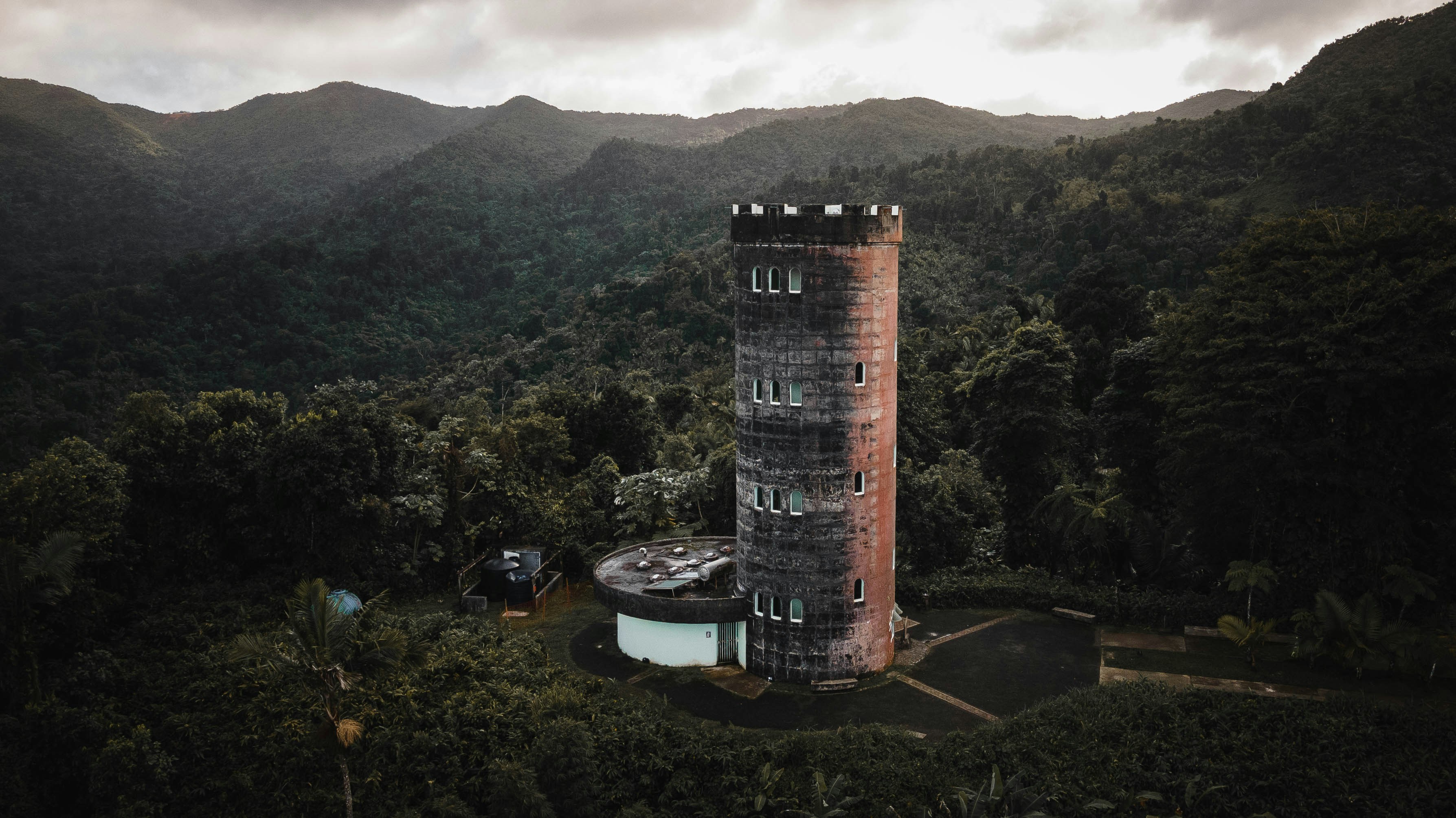 Circular tower rising above lush green landscape with distant mountains under a cloudy sky.