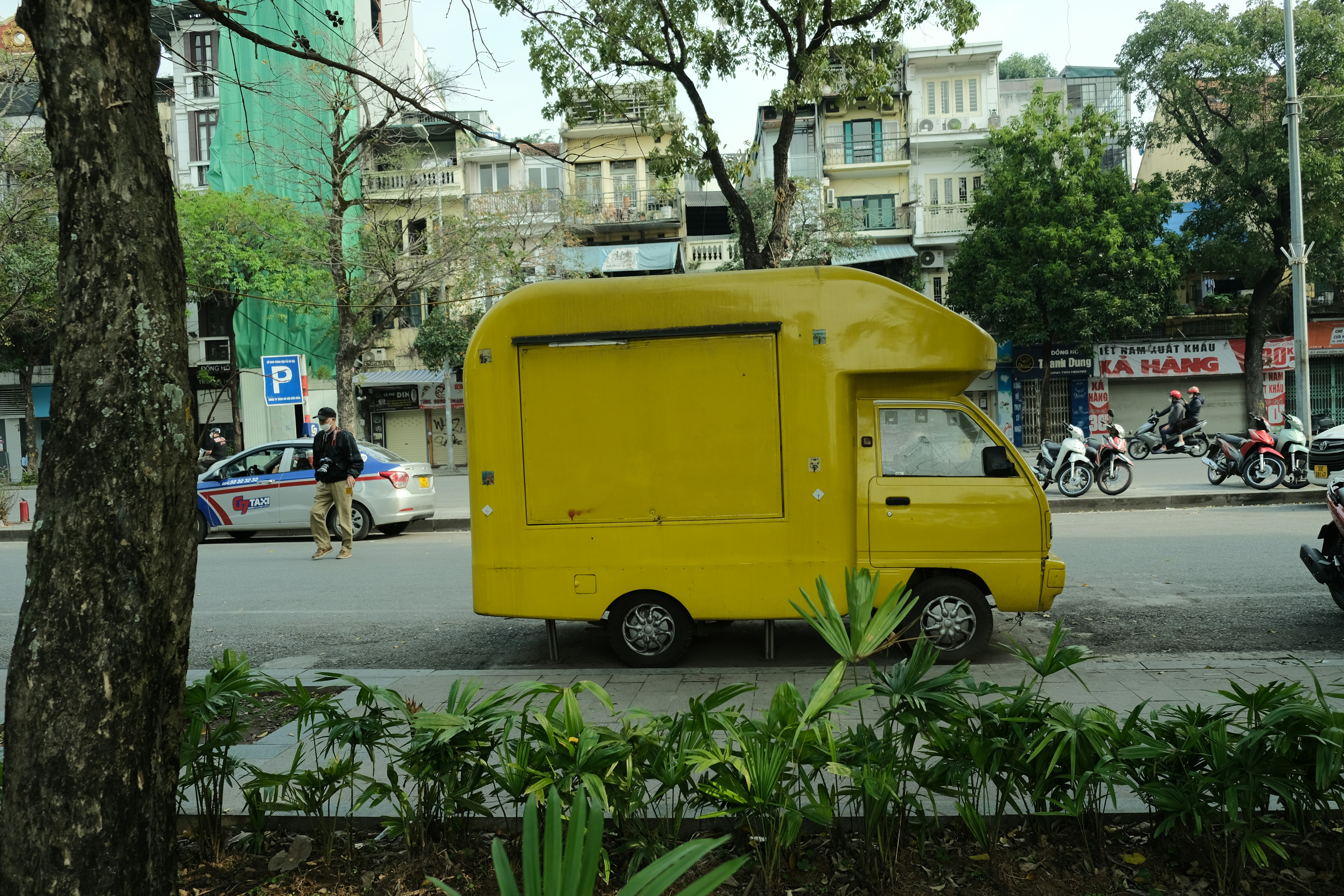 Bright yellow food truck parked along a bustling street, surrounded by urban scenery and greenery.