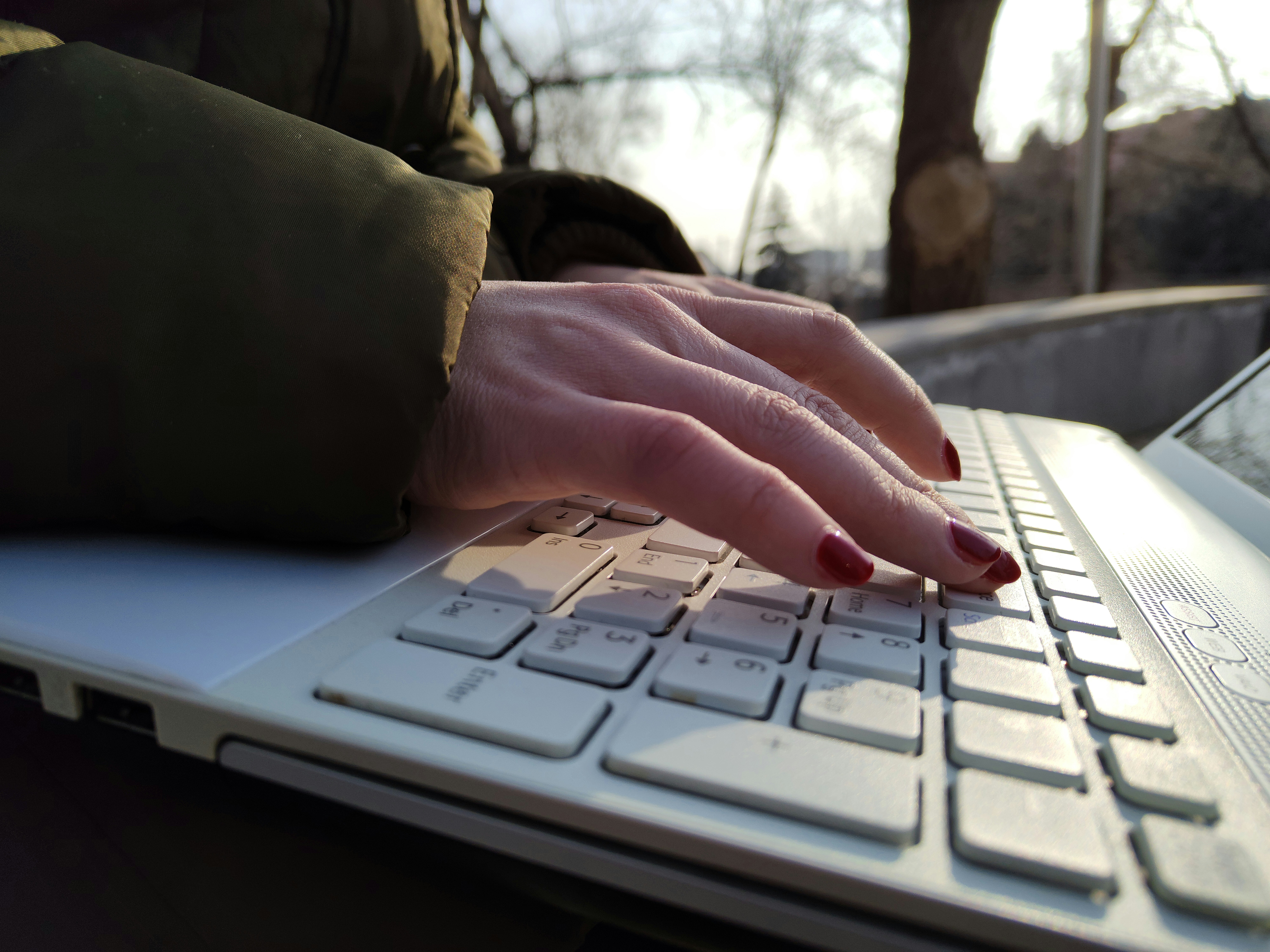 A hand with red nails gracefully typing on a laptop keyboard, surrounded by a serene outdoor setting. The soft sunlight filters through the trees, enhancing the moment.