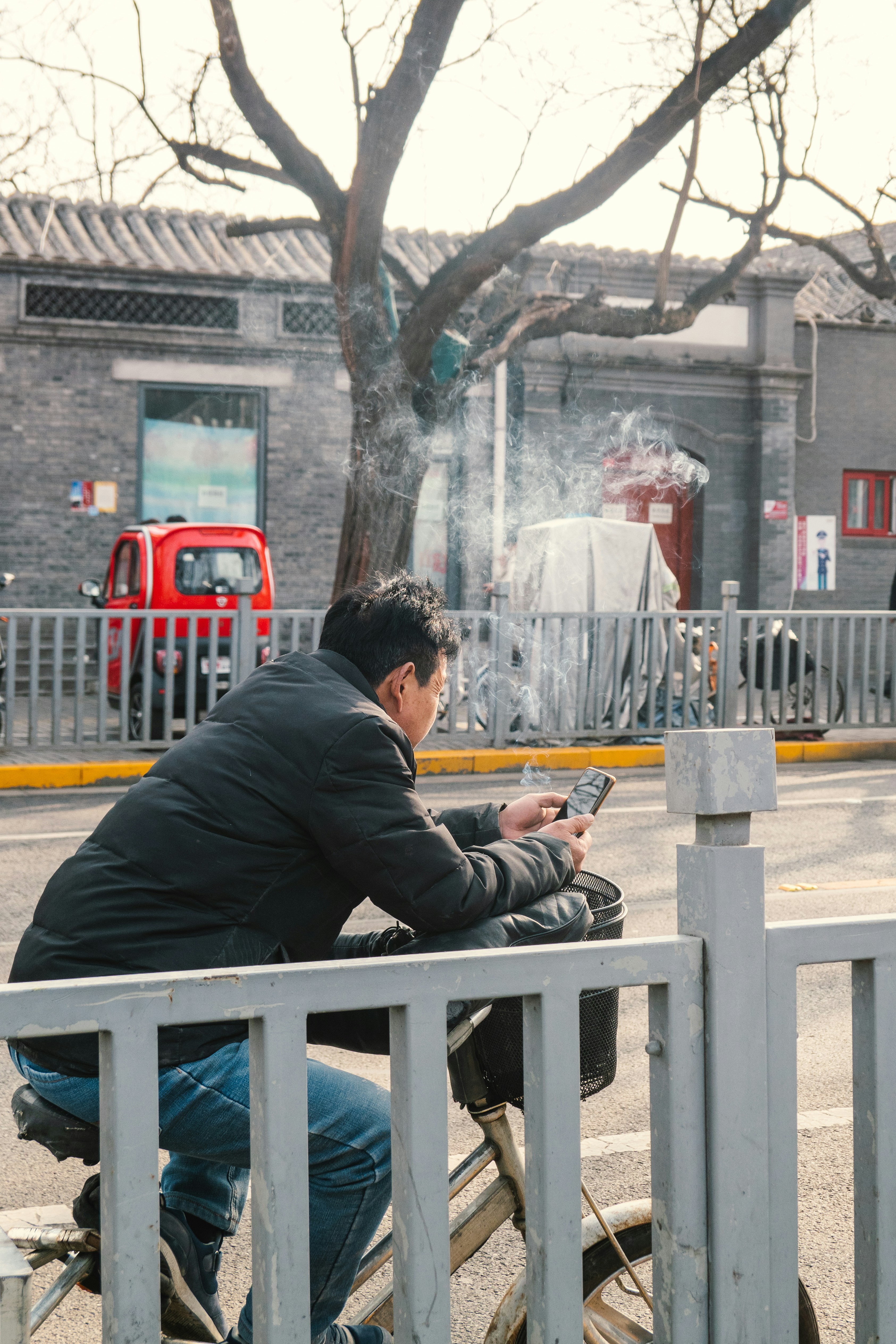 Man seated on a bicycle, engrossed in his smartphone, with a backdrop of urban architecture and a tree. Smoke wafts gently in the air.