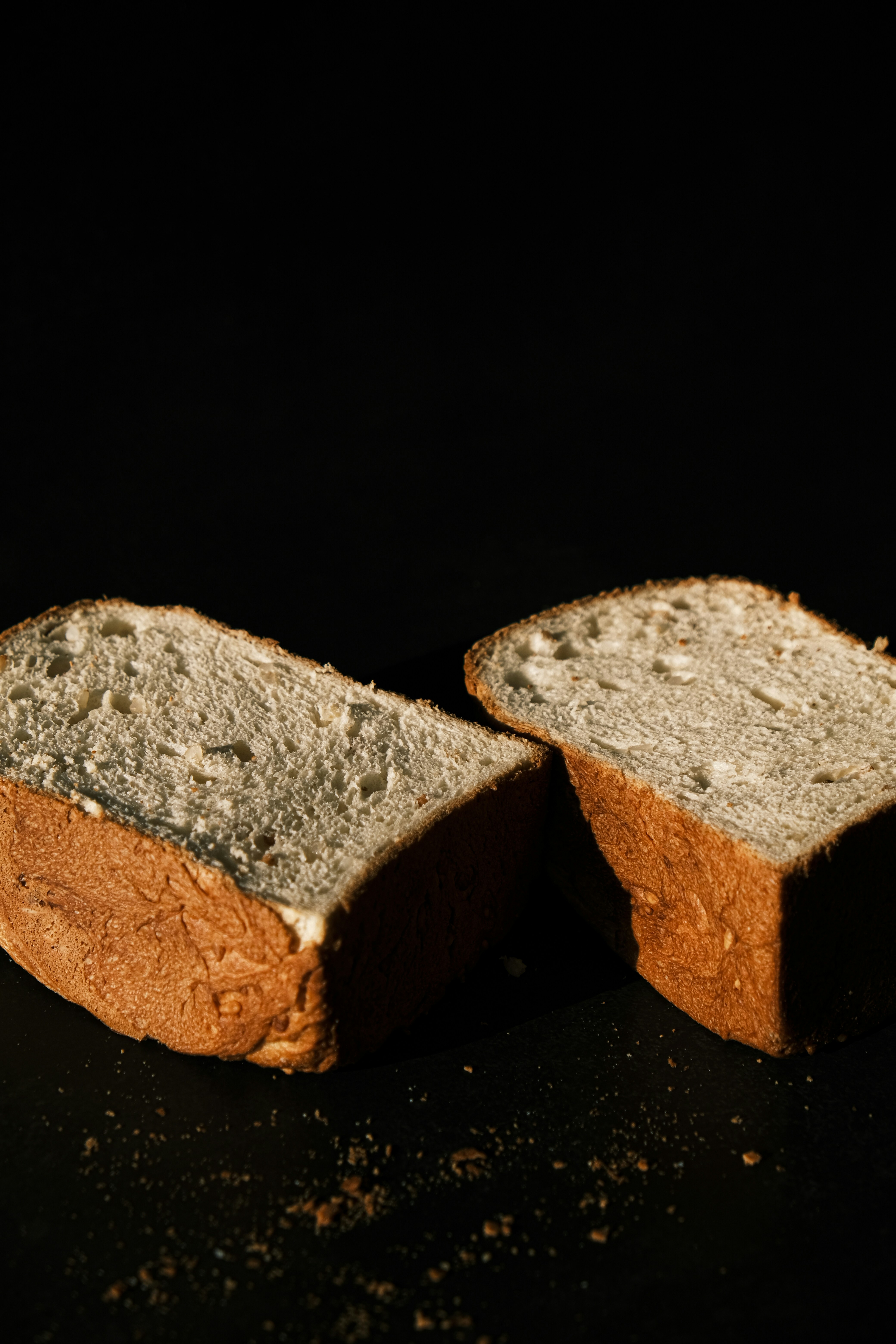 Two slices of freshly baked bread resting on a dark surface, showcasing their texture and crust. The lighting highlights the soft interior and golden-brown edges.