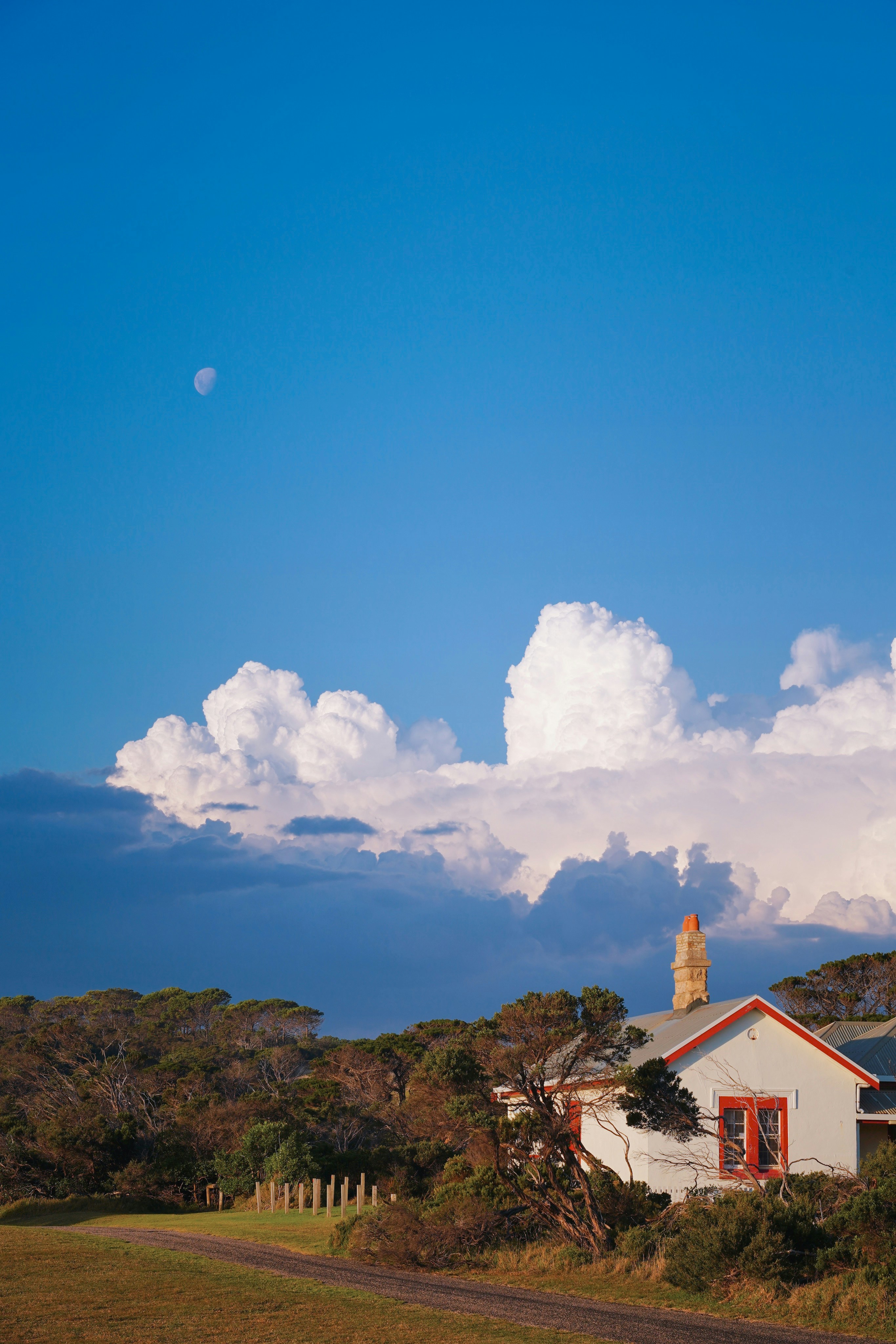 A quaint cottage nestled among lush greenery under a vibrant blue sky, with a crescent moon peeking through fluffy clouds above. 