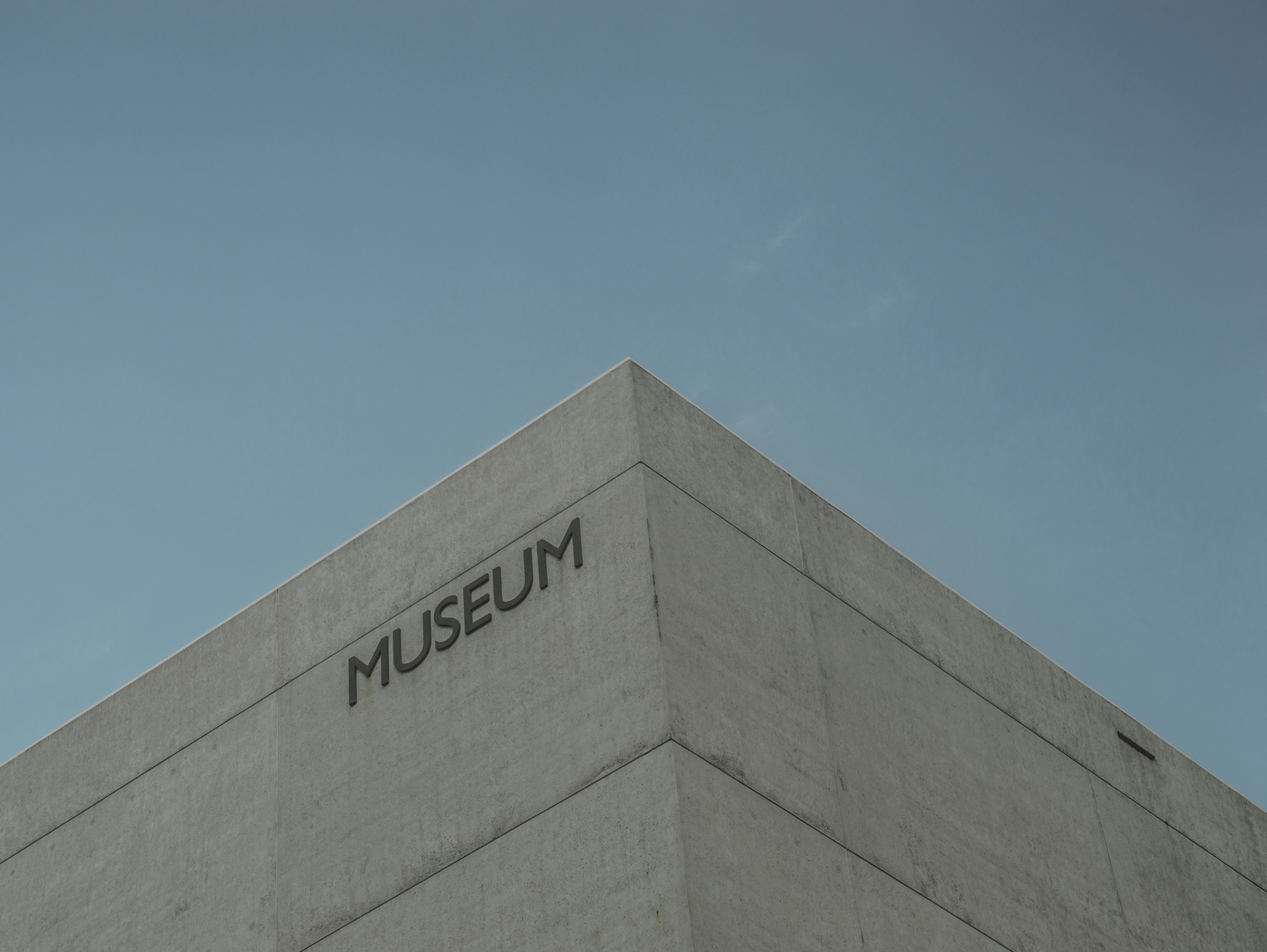A minimalist museum building features a stark concrete exterior with the word 'MUSEUM' prominently displayed against a clear blue sky.
