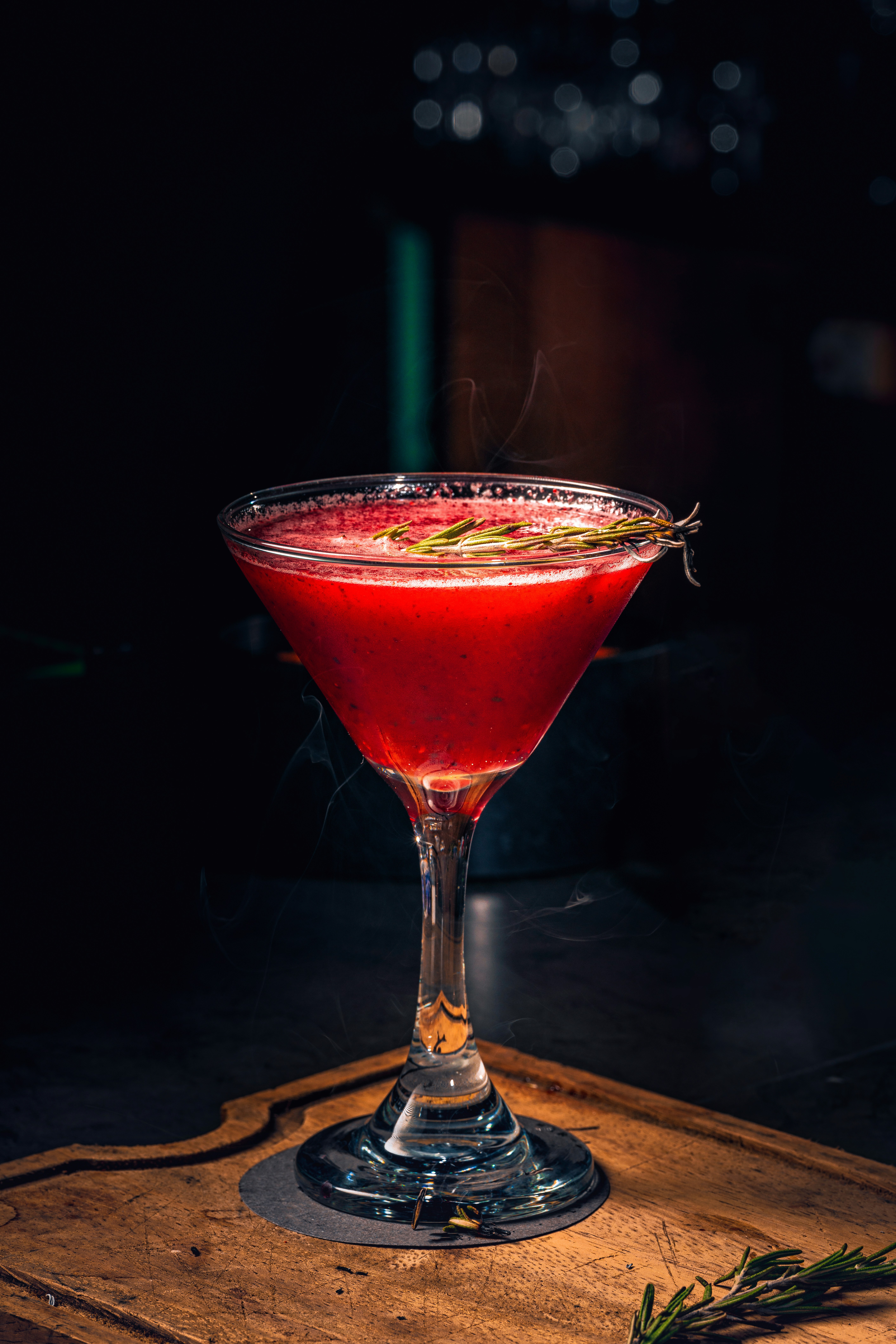 A vibrant red cocktail garnished with rosemary, resting on a wooden board with soft smoke wafting above it.