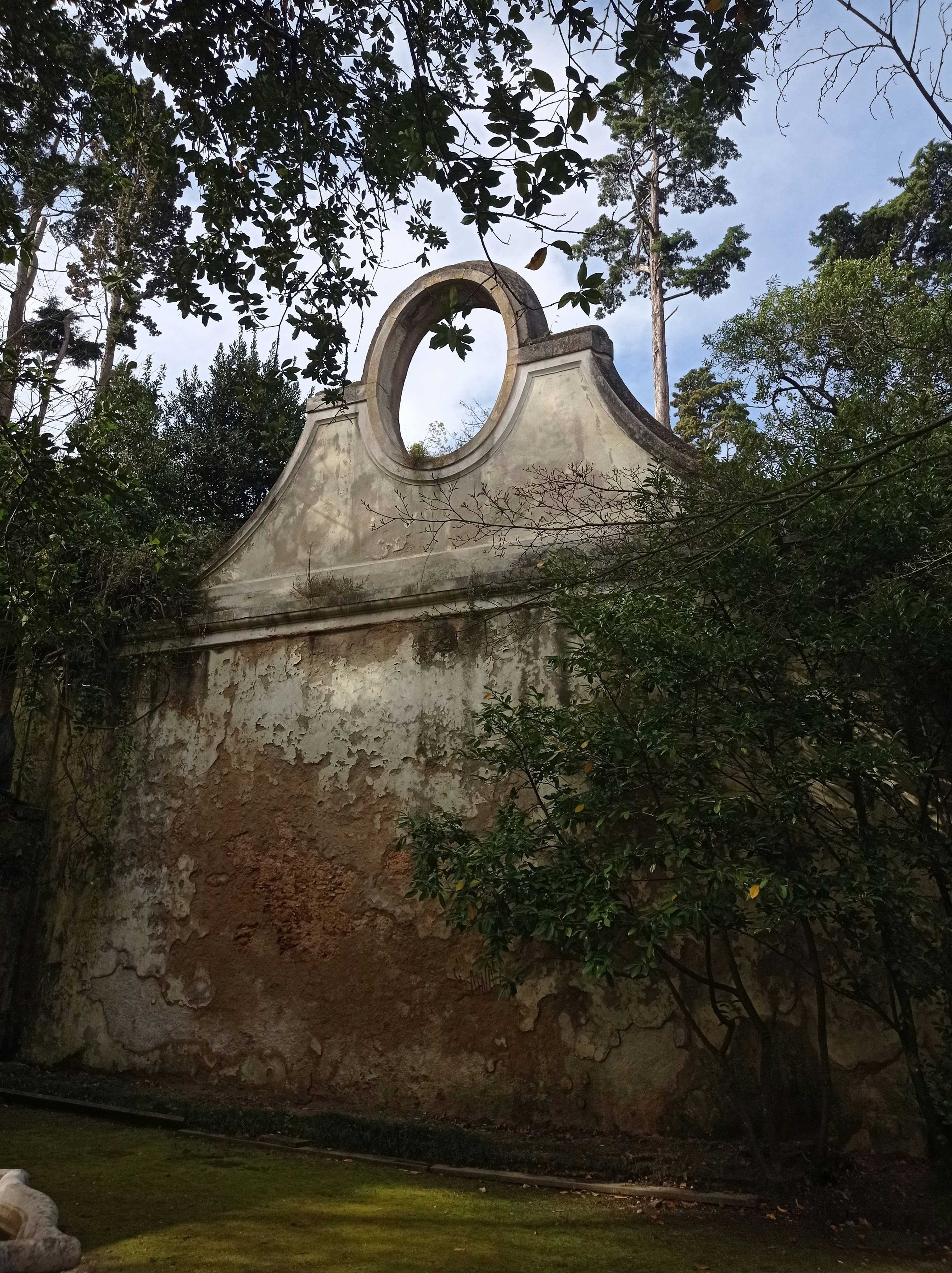 A weathered wall adorned with a circular arch, surrounded by lush greenery in a serene setting. Nature intertwines with the remnants of architecture.
