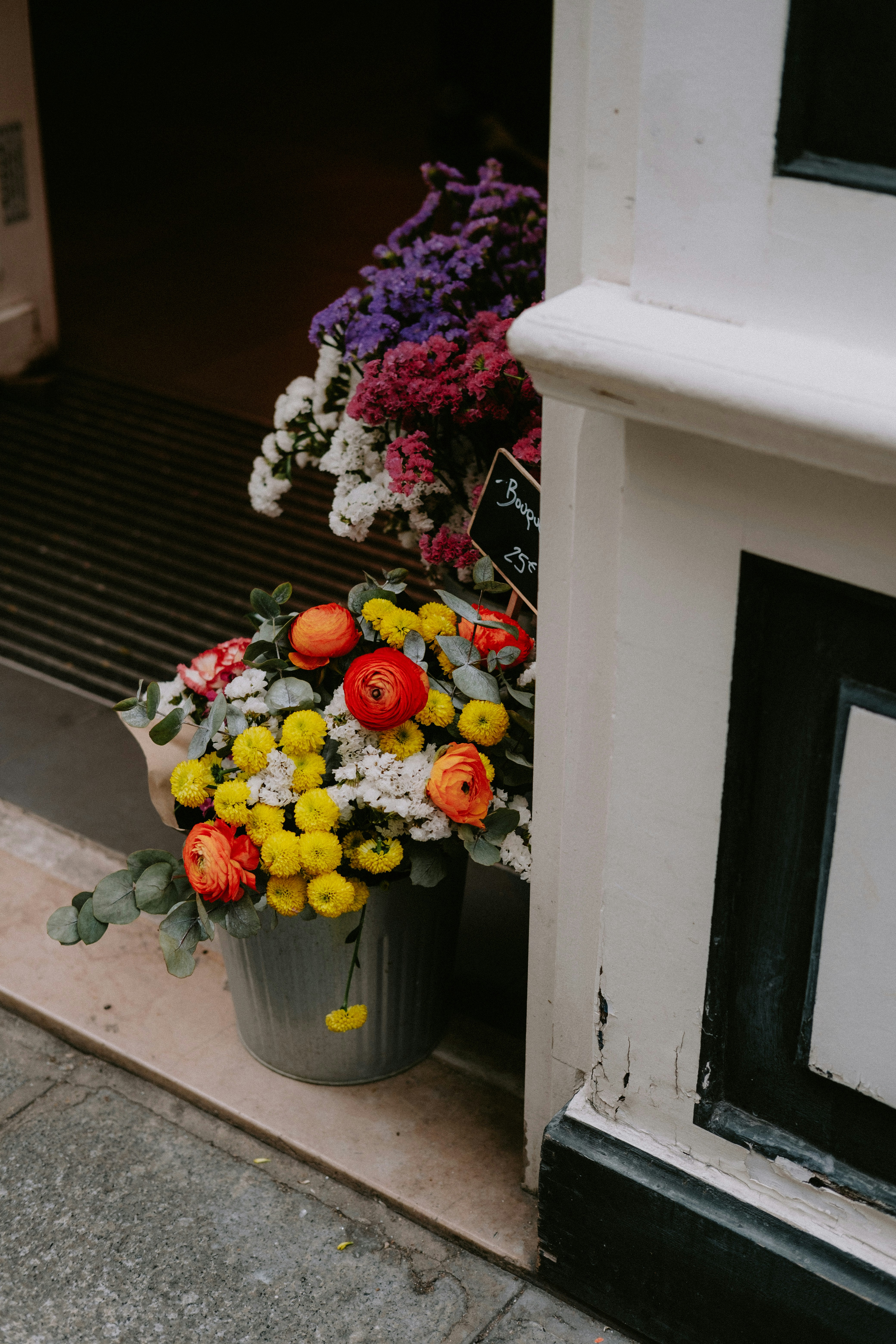 Vibrant bouquet of flowers in a metal bucket near a doorway, featuring orange ranunculus and yellow chrysanthemums. A small chalkboard sign adds a charming touch.