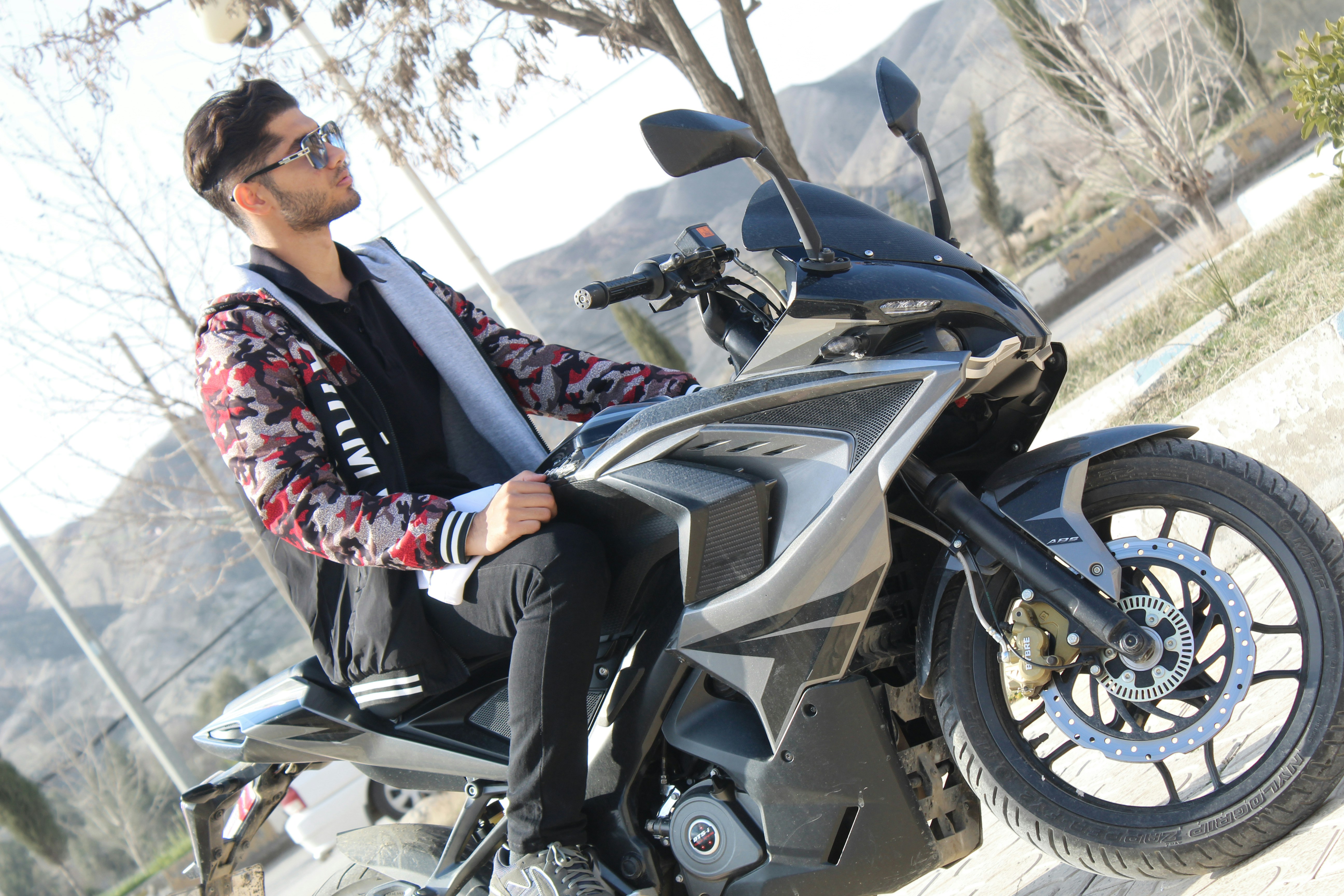 Young man dressed in a stylish jacket sitting on a sleek motorcycle, surrounded by a scenic backdrop of mountains and trees.