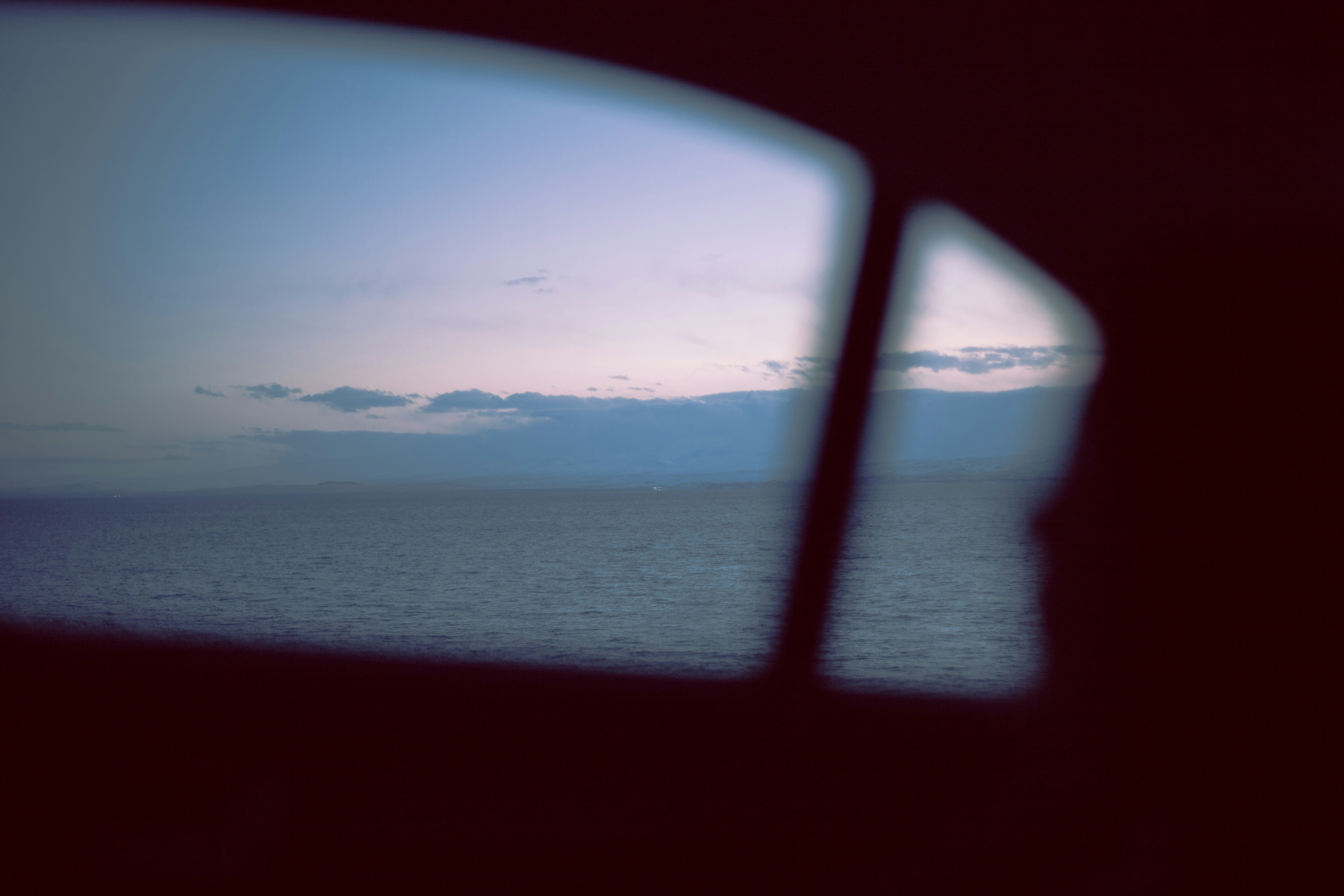 View of the ocean through a car window at dusk, with soft colors blending in the sky. The silhouette of a figure is visible in the foreground.