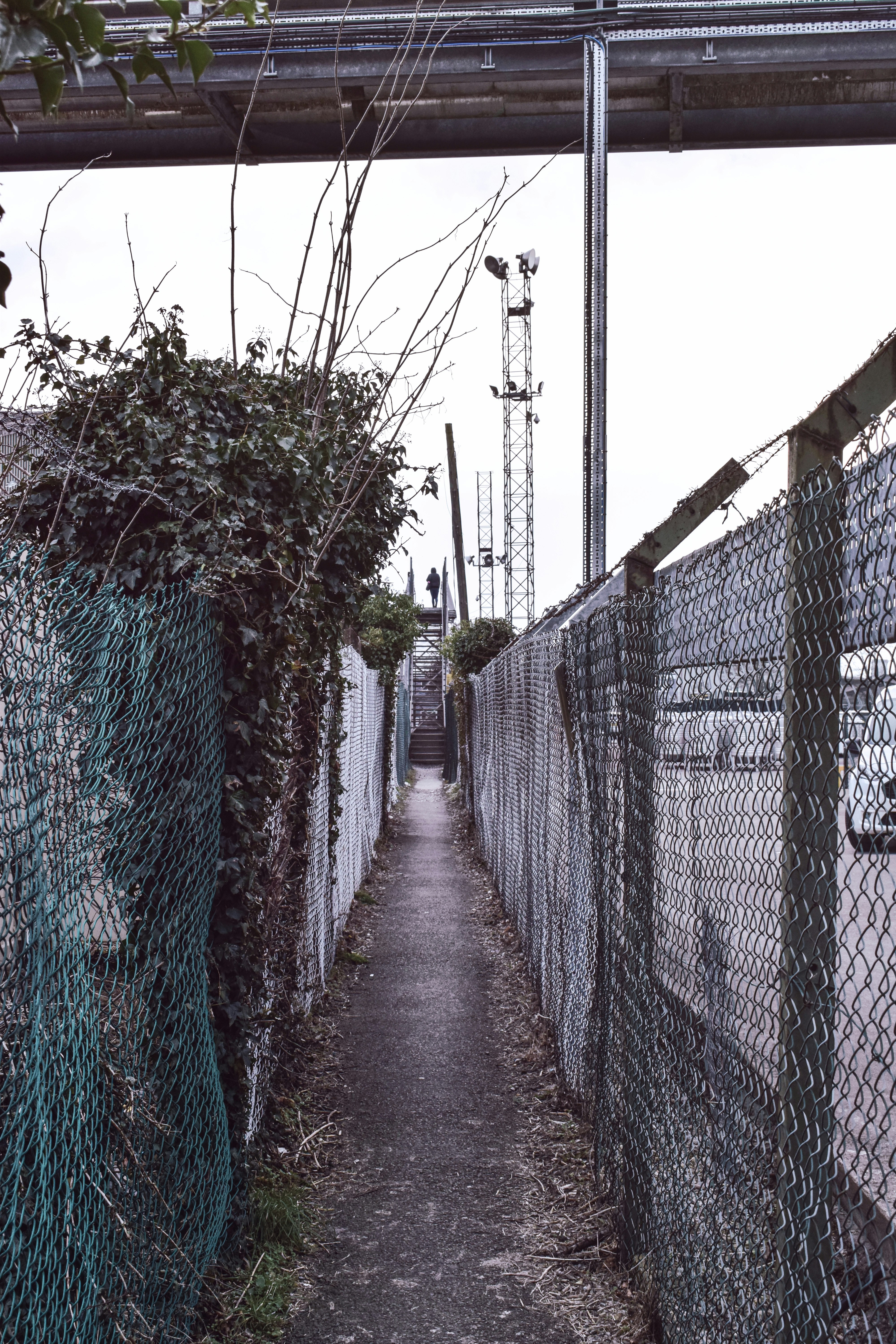 Narrow pathway flanked by chain-link fences and industrial structures under an overcast sky.