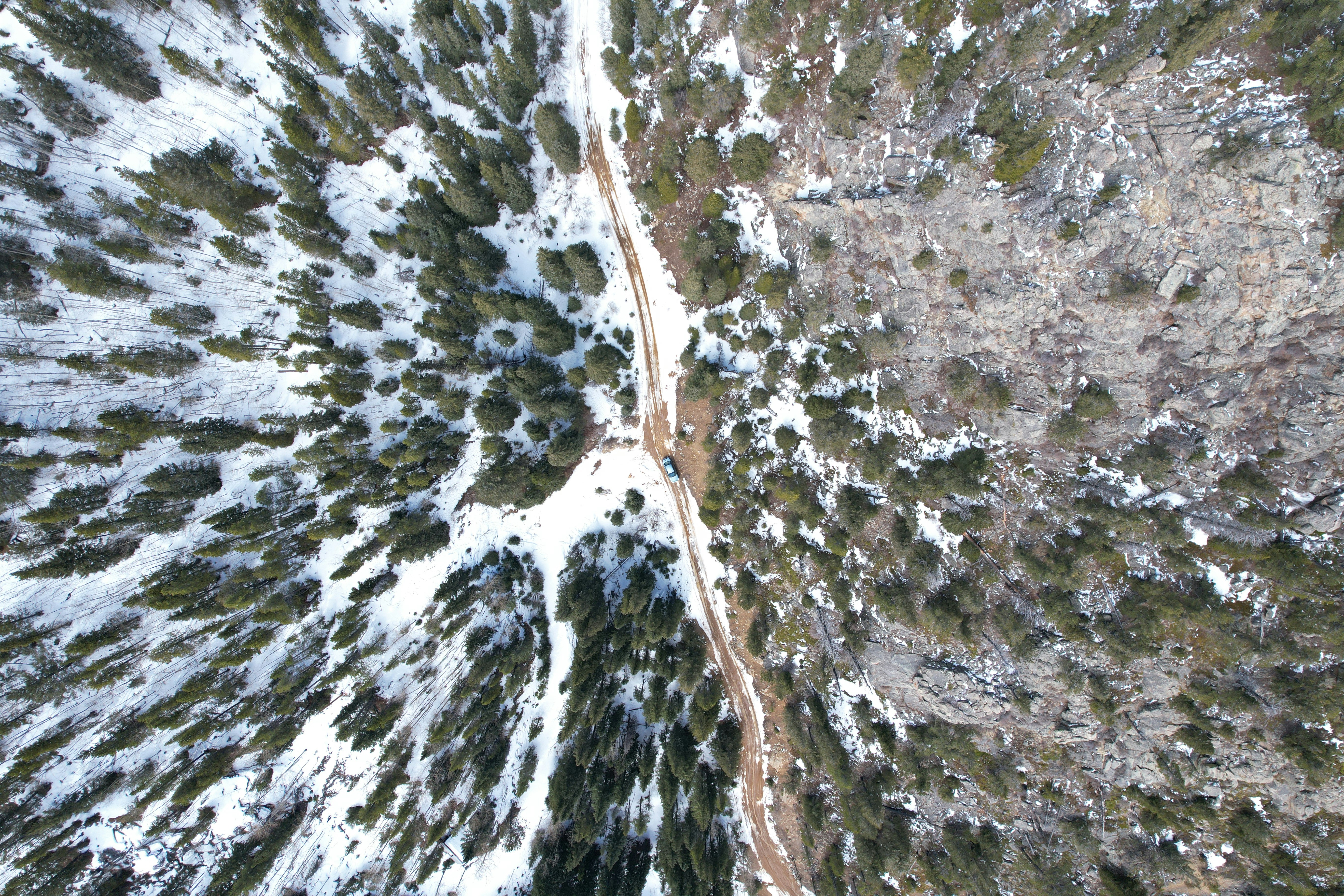 Aerial view showcasing a winding dirt path through a snowy landscape surrounded by evergreen trees and rocky terrain.