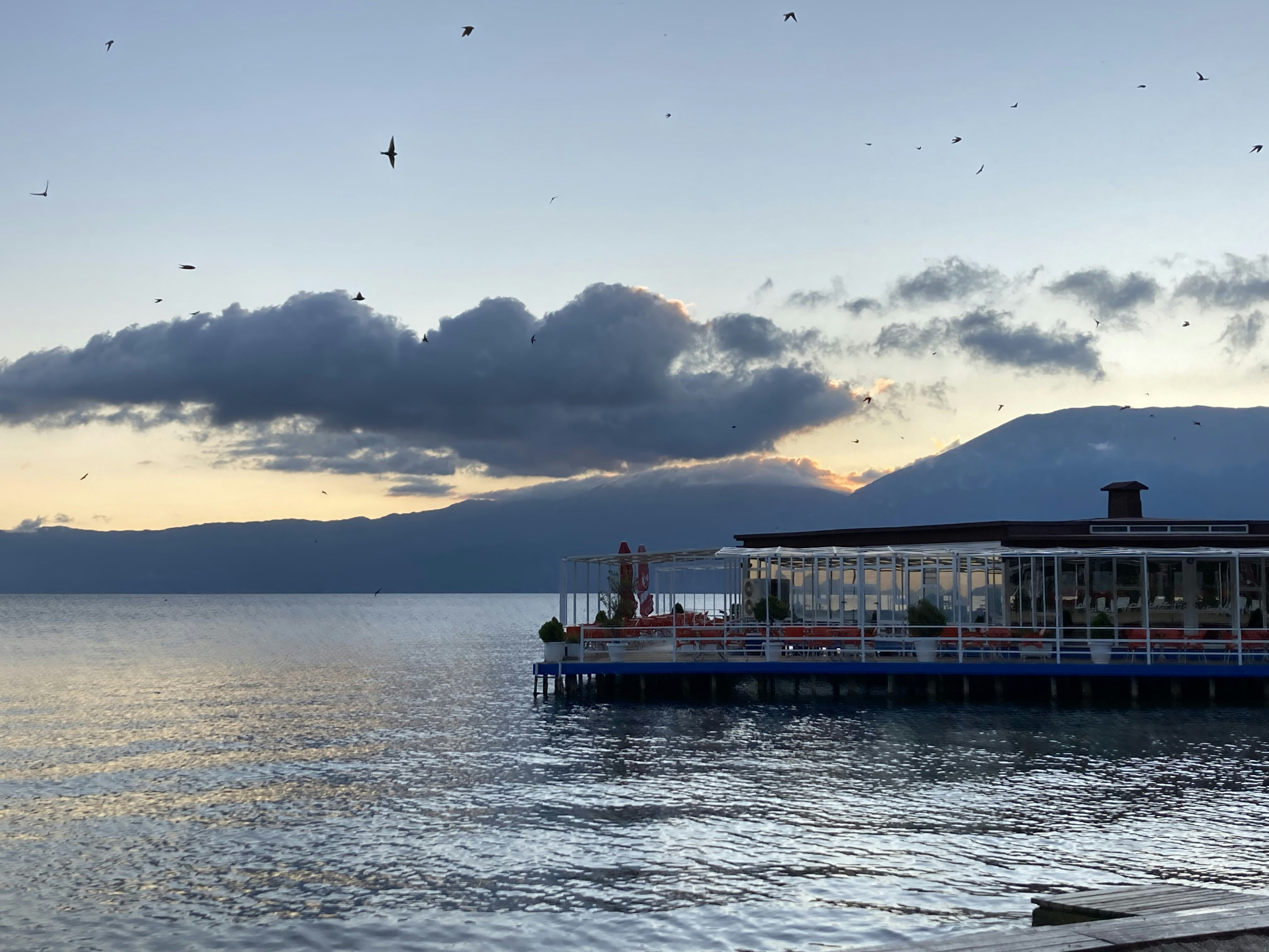 Lakeside restaurant with a wooden deck reflecting the evening sky, surrounded by distant mountains and flying birds.