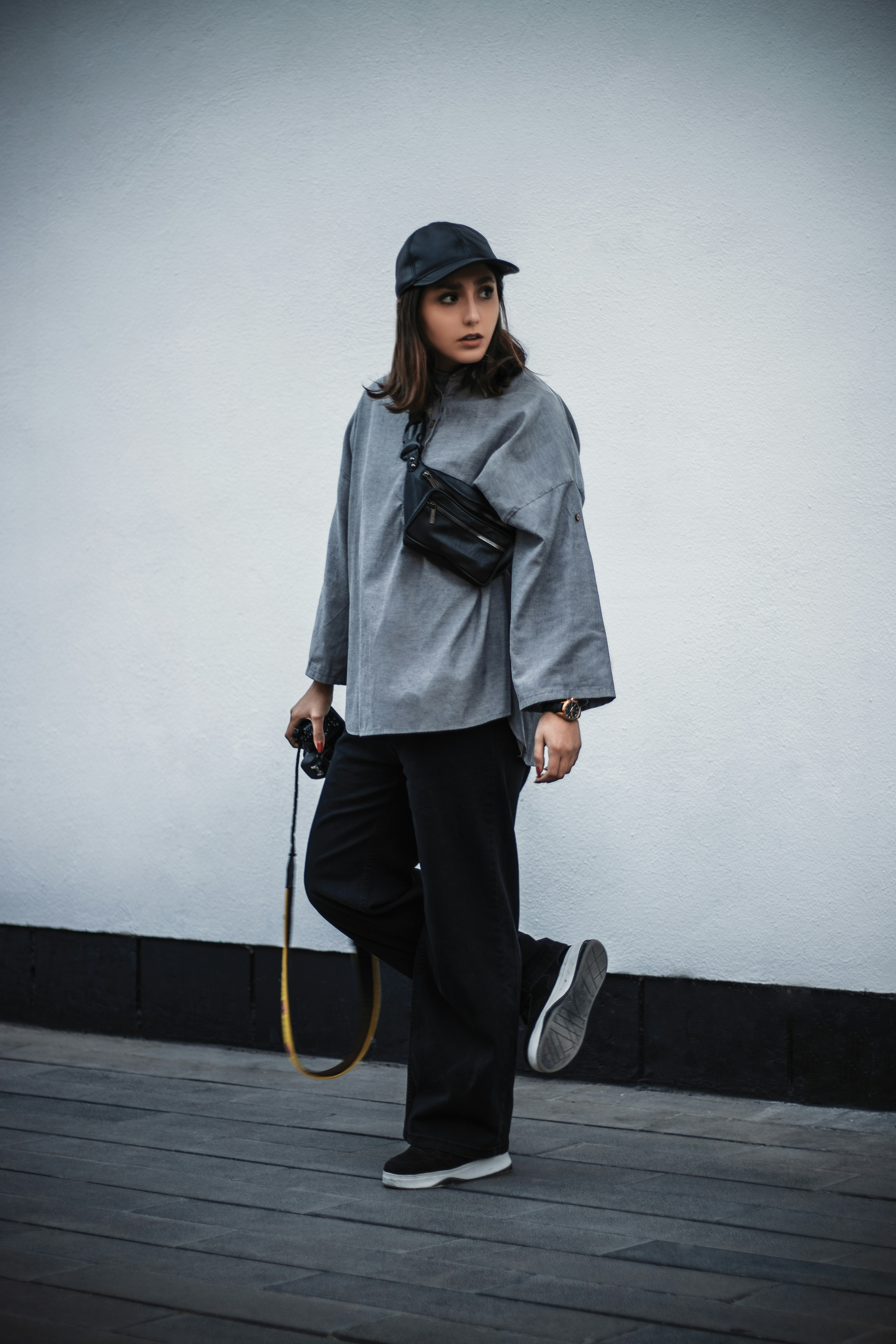 A young woman in a stylish gray jacket and cap stands against a minimalist wall, holding a camera and exuding an air of urban sophistication.