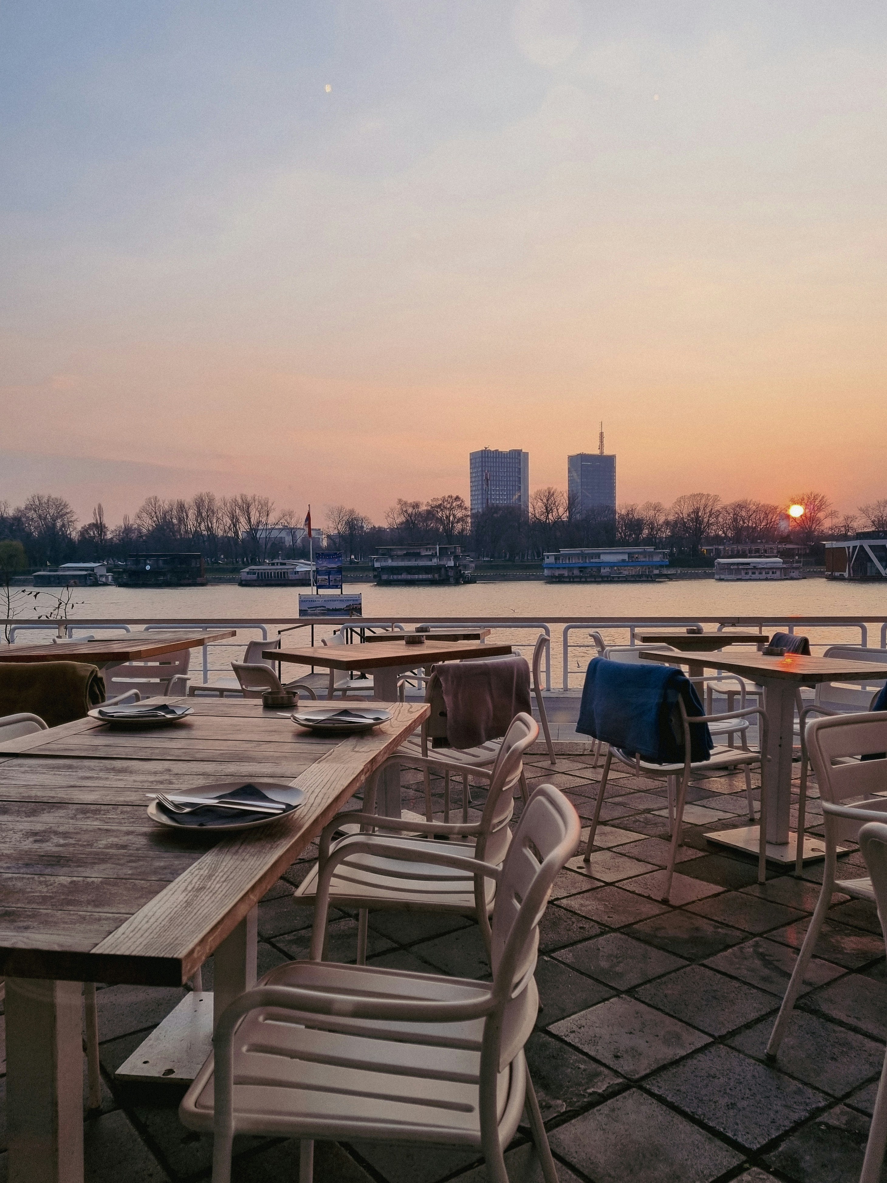 Tables set for dining on a riverside terrace with a sunset backdrop illuminating the skyline. The scene evokes a sense of tranquility and anticipation for the evening ahead.