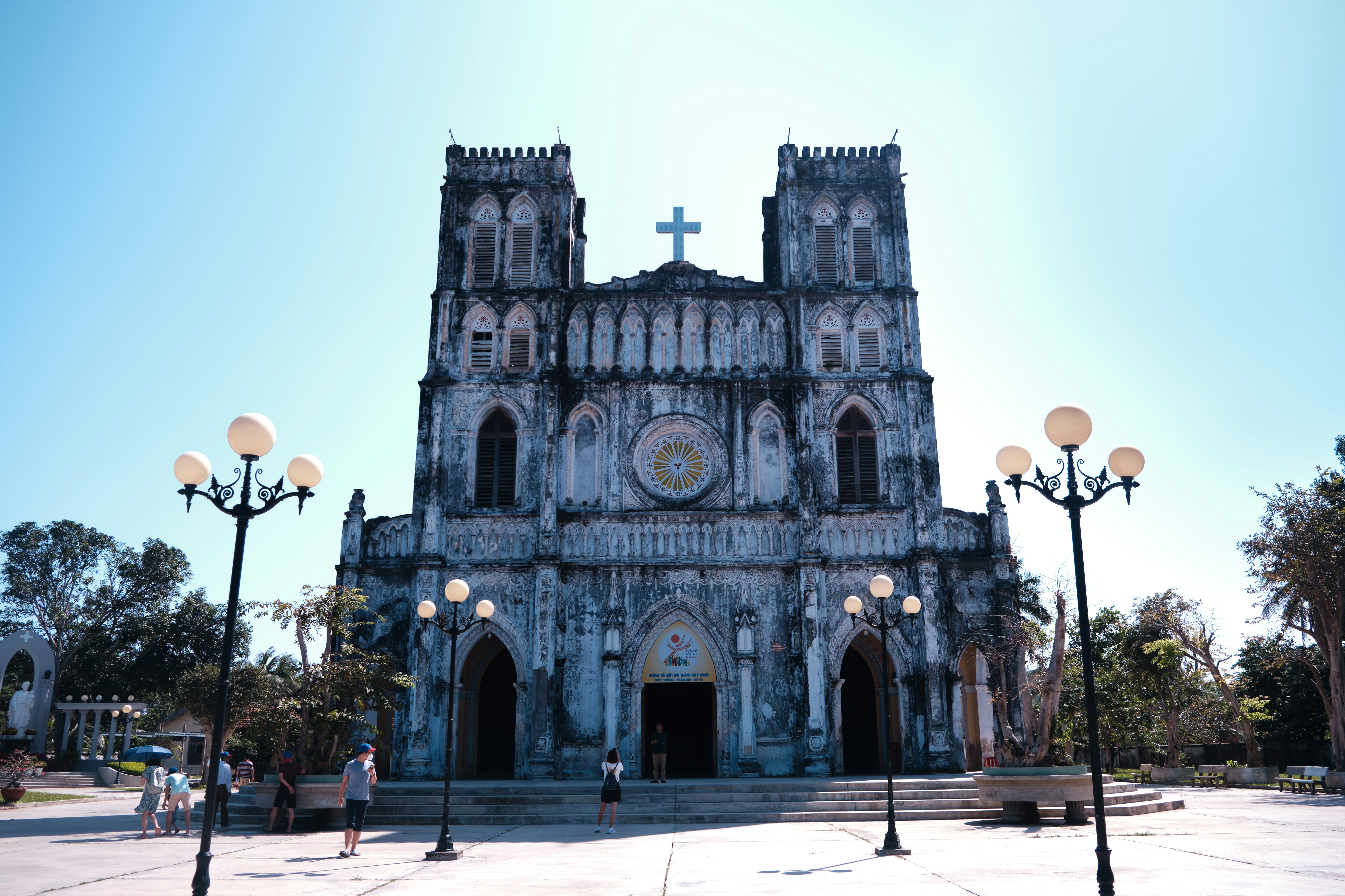 Historic church with intricate architecture and a prominent cross, surrounded by lampposts and visitors enjoying the sunny day.