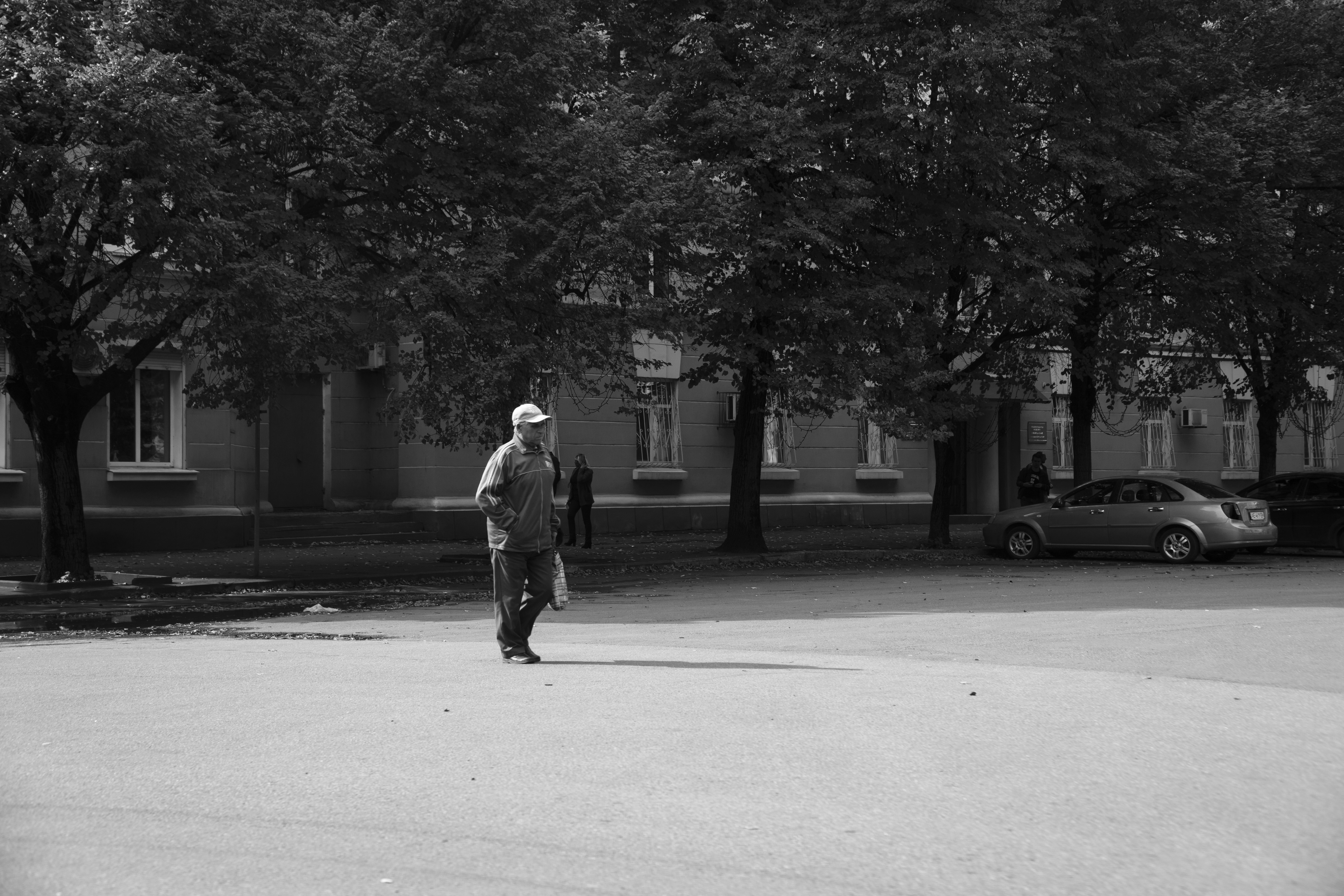 An elderly man walks across a deserted street, framed by trees and distant figures, evoking a sense of quiet reflection. Black and white aesthetic enhances the mood.