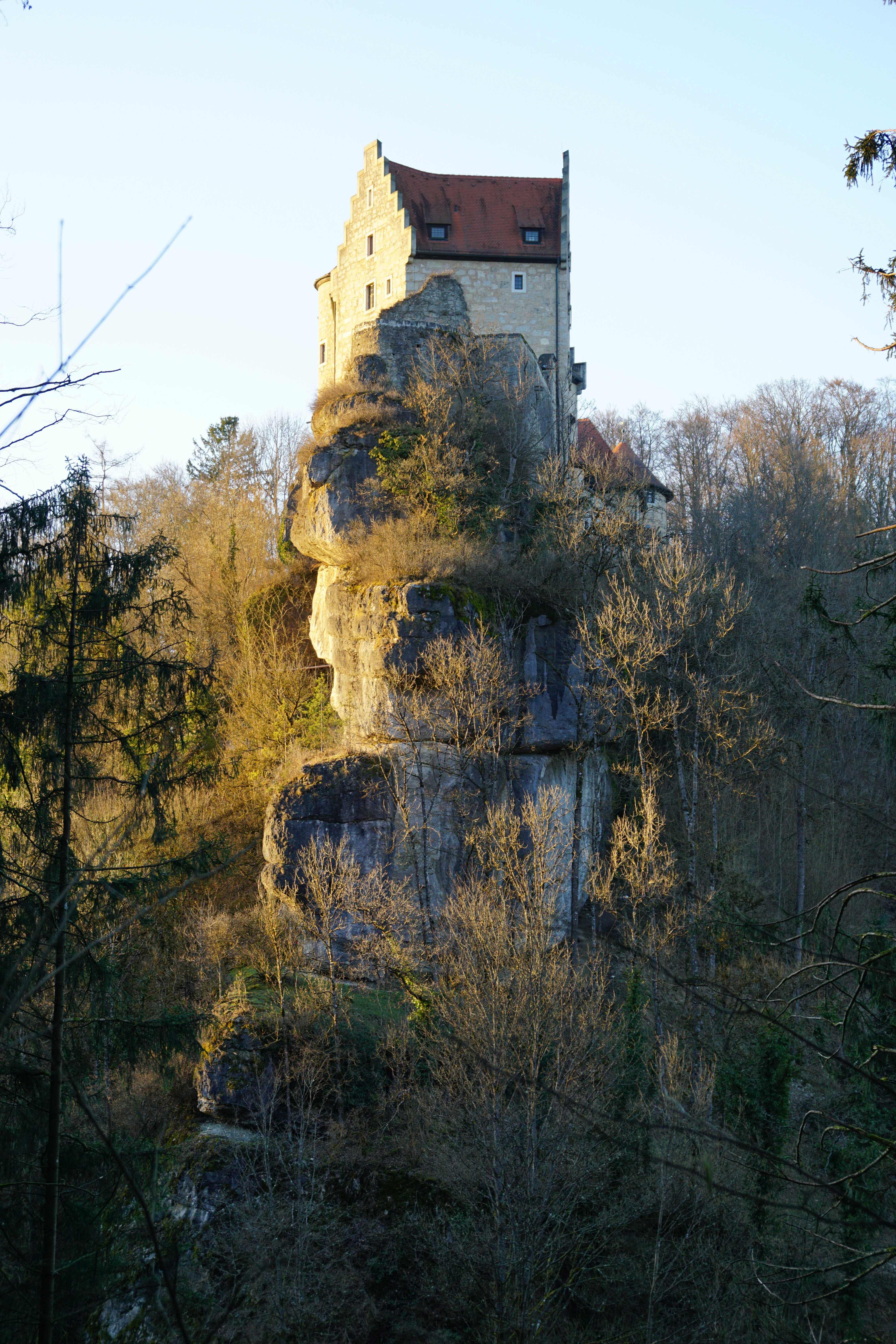 A historic castle perched atop a rocky outcrop, surrounded by trees in a serene landscape. The structure showcases unique architecture against a backdrop of soft evening light.
