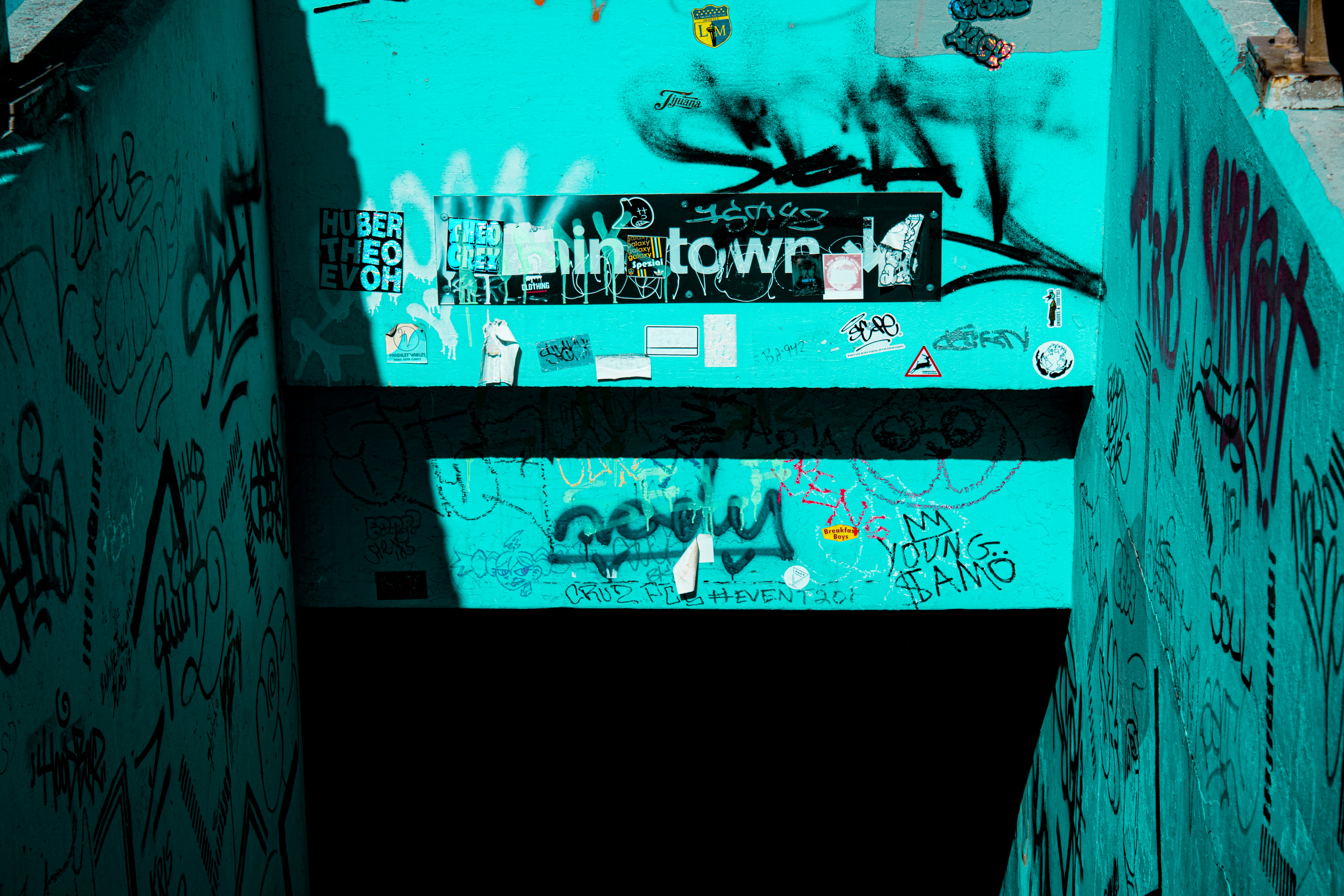 Vibrant turquoise tunnel adorned with eclectic graffiti and stickers, leading into shadowy depths. The artwork reflects urban creativity and expression.
