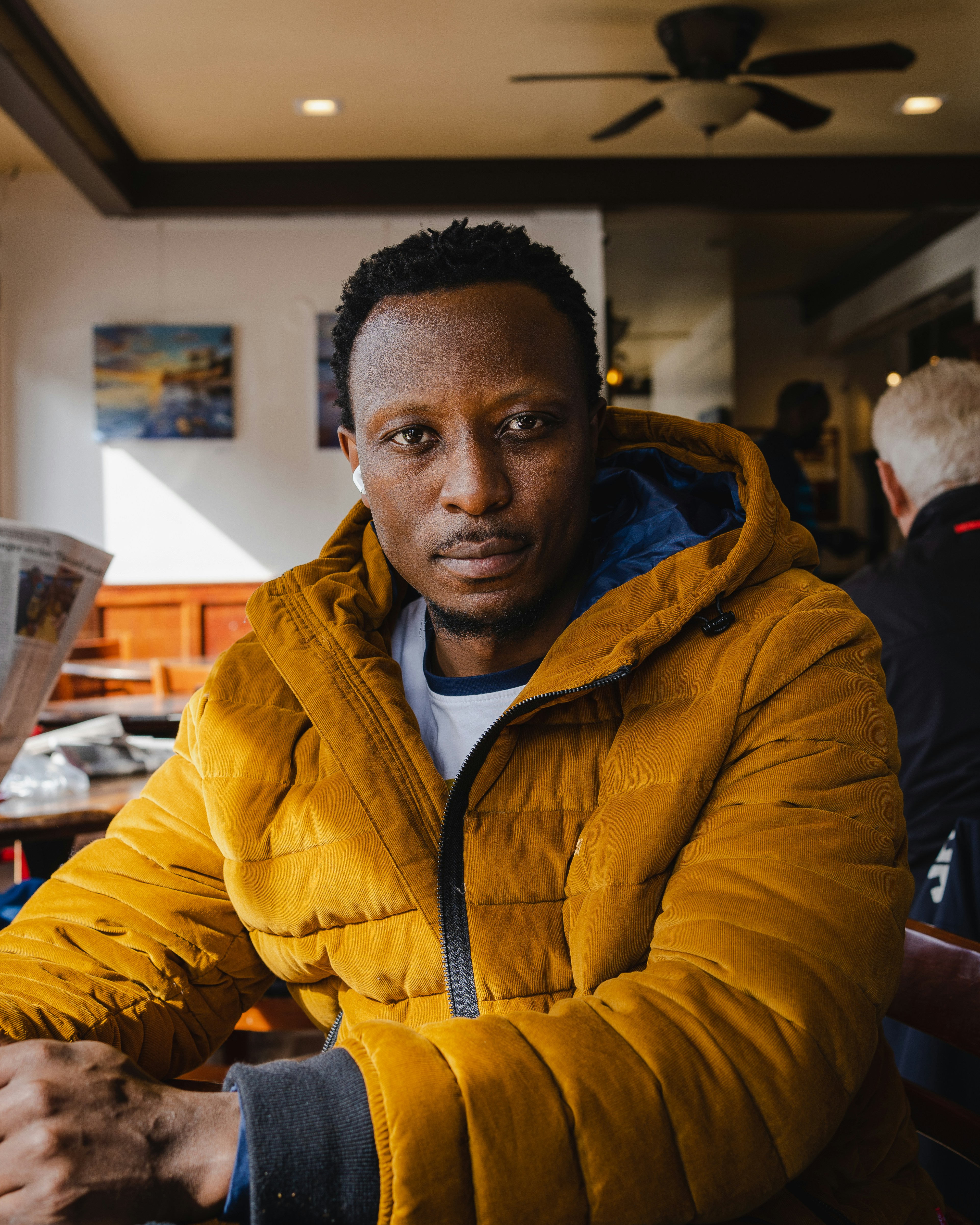 Individual in a yellow jacket seated at a café table, gazing thoughtfully while sunlight filters through the window.