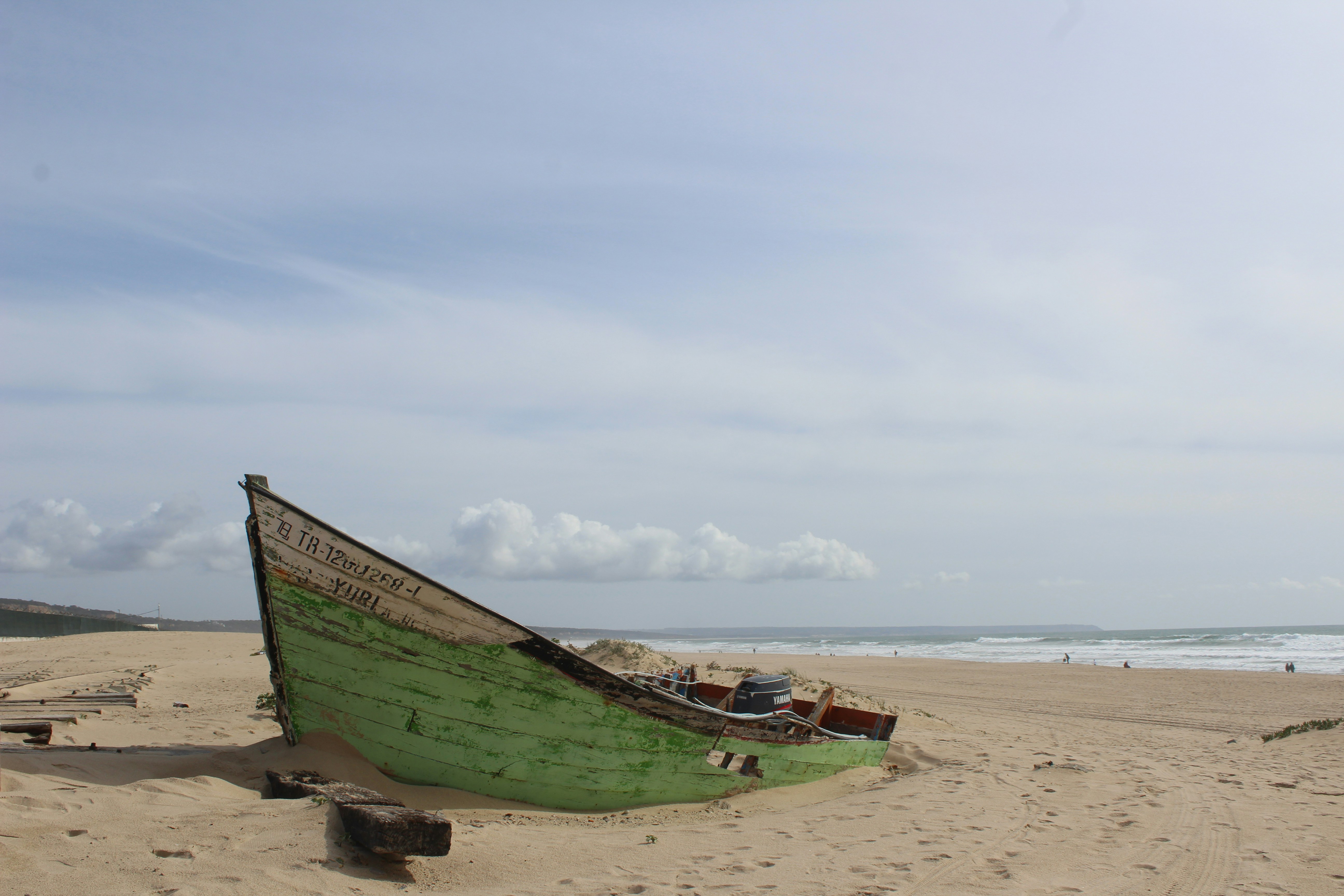 Weathered fishing boat resting on a sandy beach, surrounded by gentle waves and distant clouds.