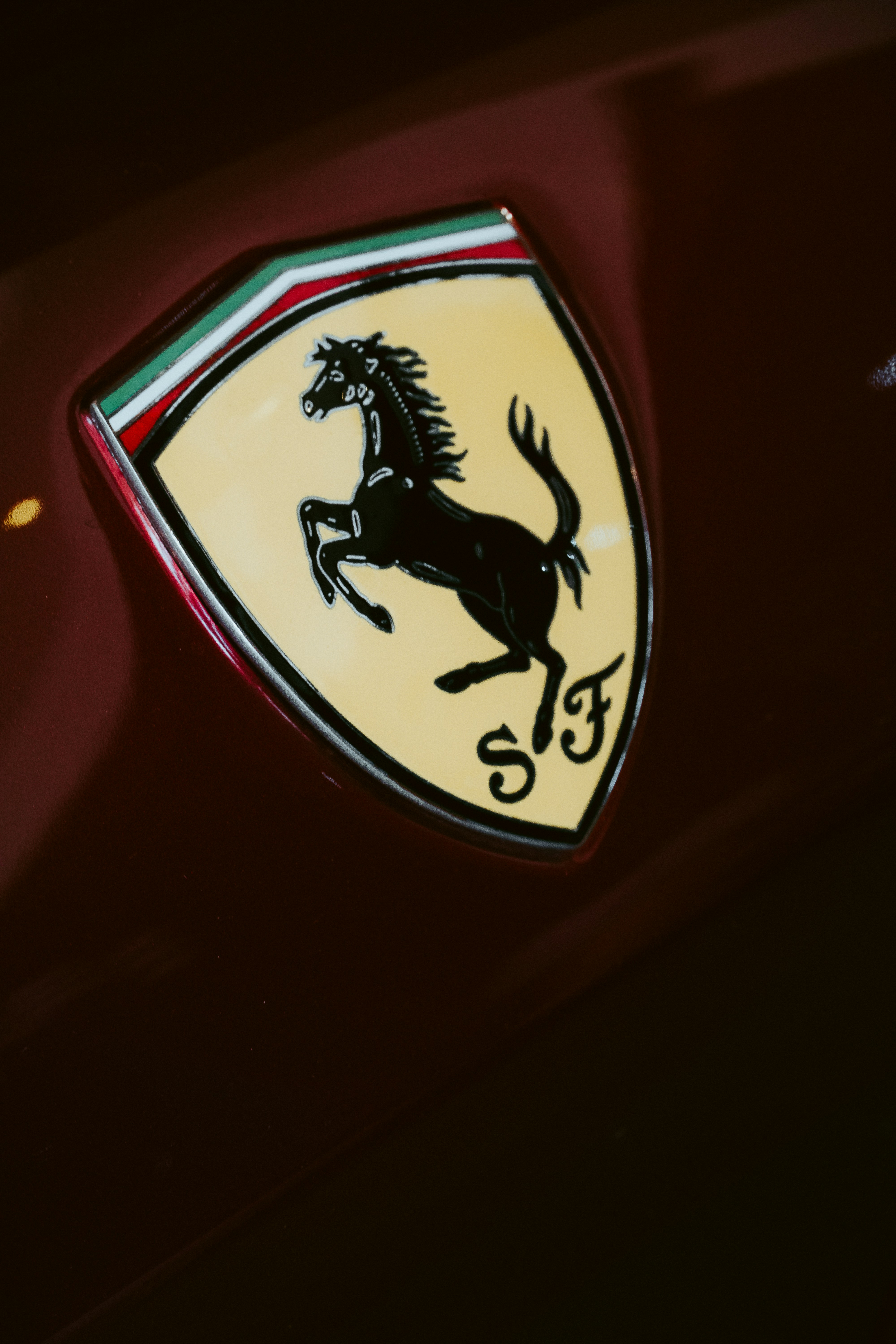 Ferrari logo featuring a prancing horse against a cream background, framed by a shield with green, white, and red stripes. The emblem represents luxury and high performance.