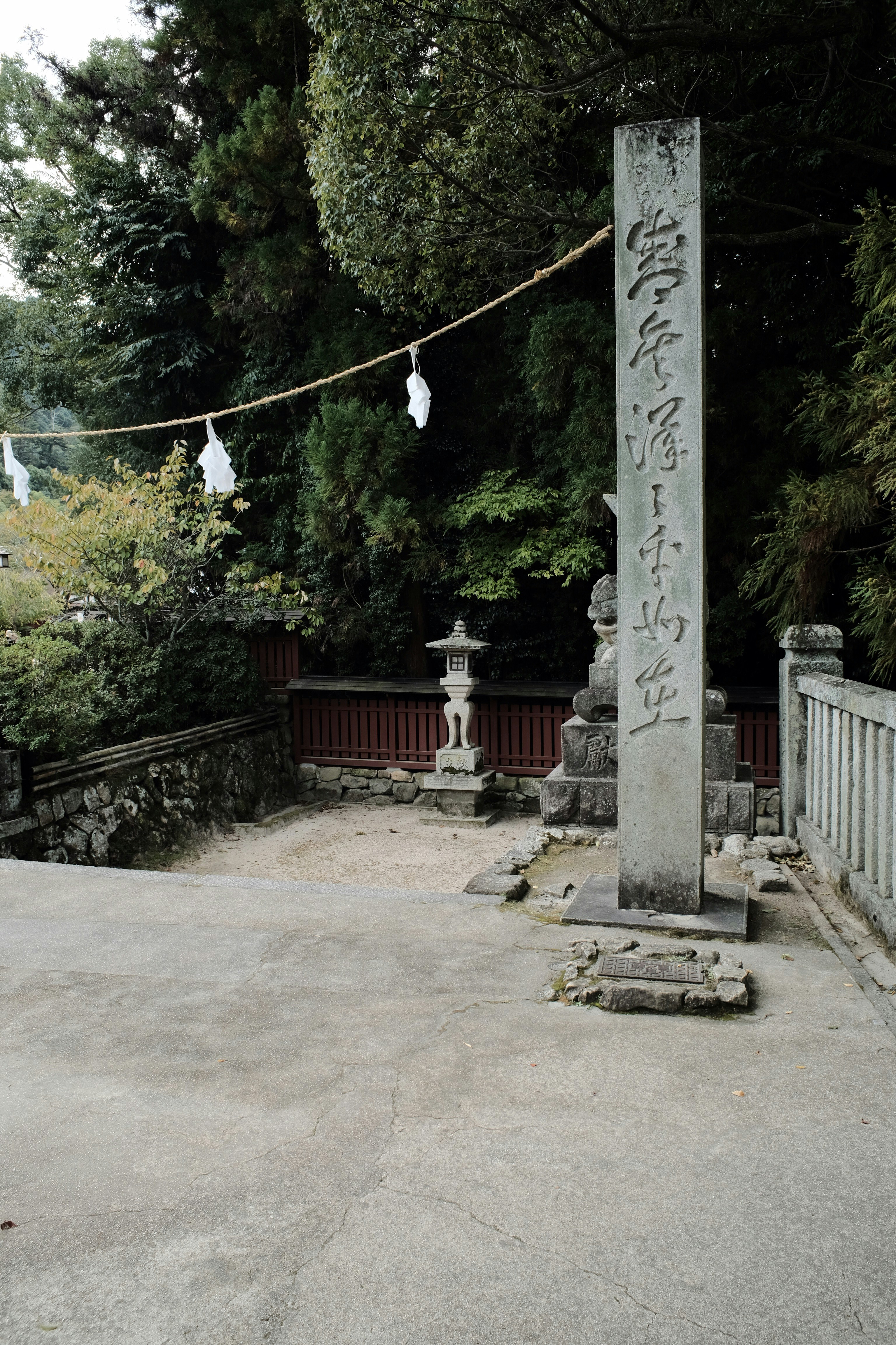 A weathered stone monument inscribed with characters stands at the entrance of a tranquil temple, surrounded by lush greenery and a lantern in the background.