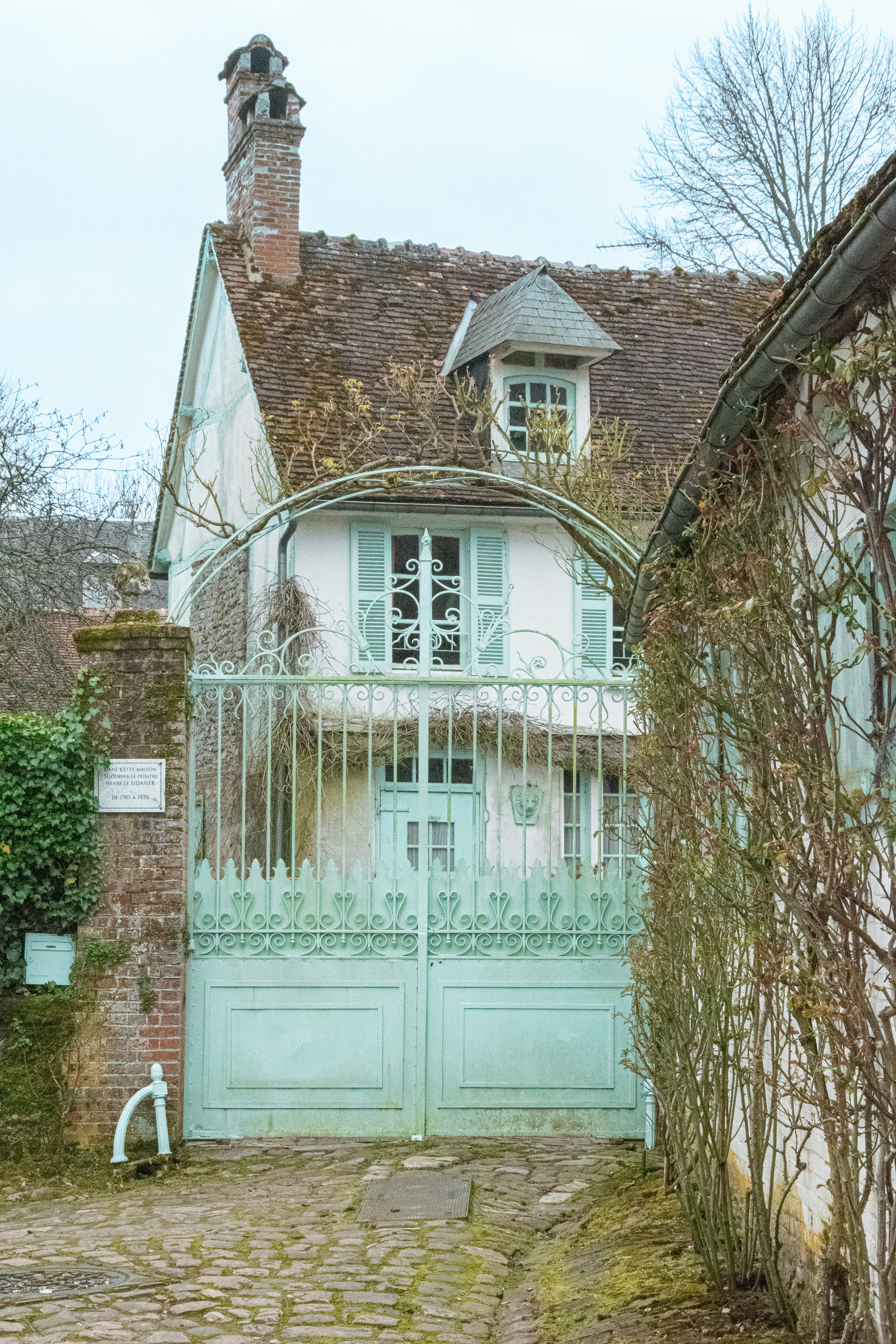 A charming mint-green gate leads to a quaint house, framed by lush greenery and cobblestone path. The architecture reflects a serene, rustic elegance.