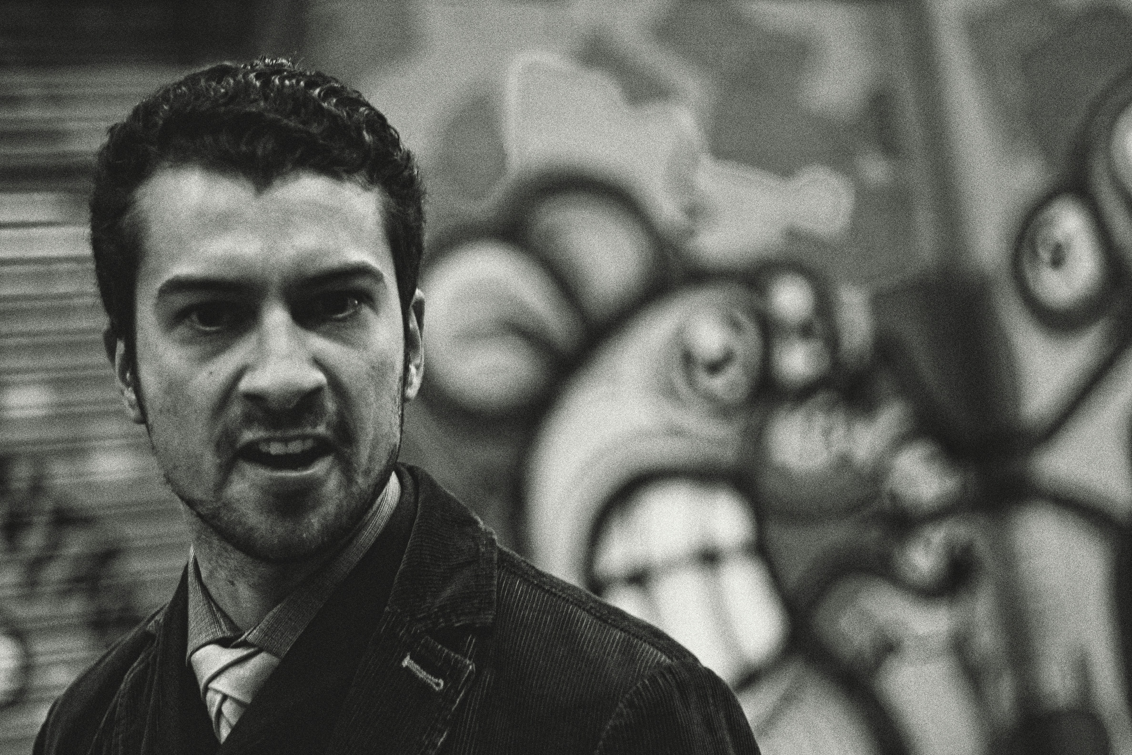 Man in a suit with an intense expression stands in front of a vibrant graffiti mural.