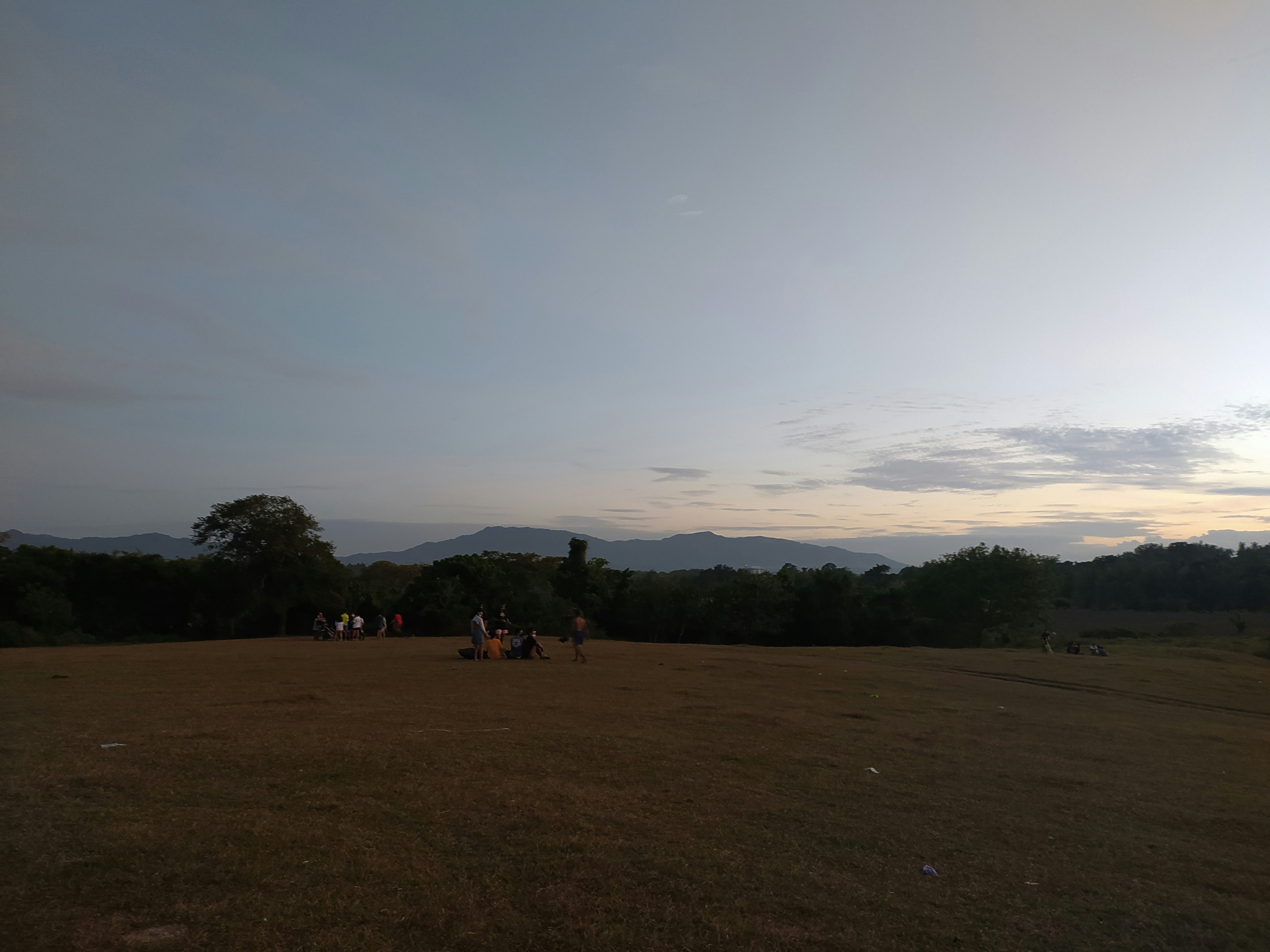 A serene landscape at dusk, featuring a group of people enjoying a gathering on a grassy field with mountains in the background.