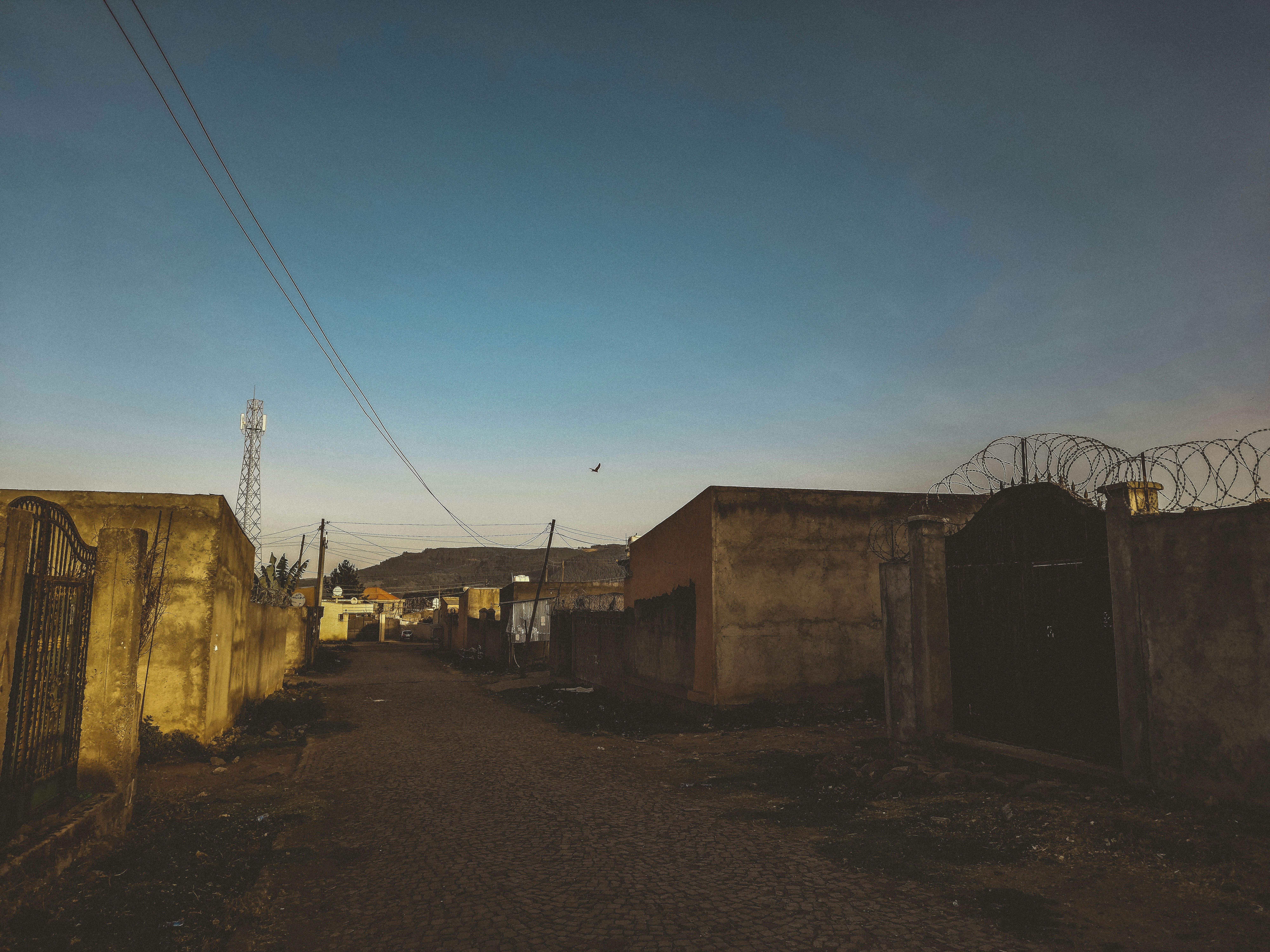 Narrow pathway flanked by weathered walls and barbed wire, leading towards distant hills under a soft blue sky.