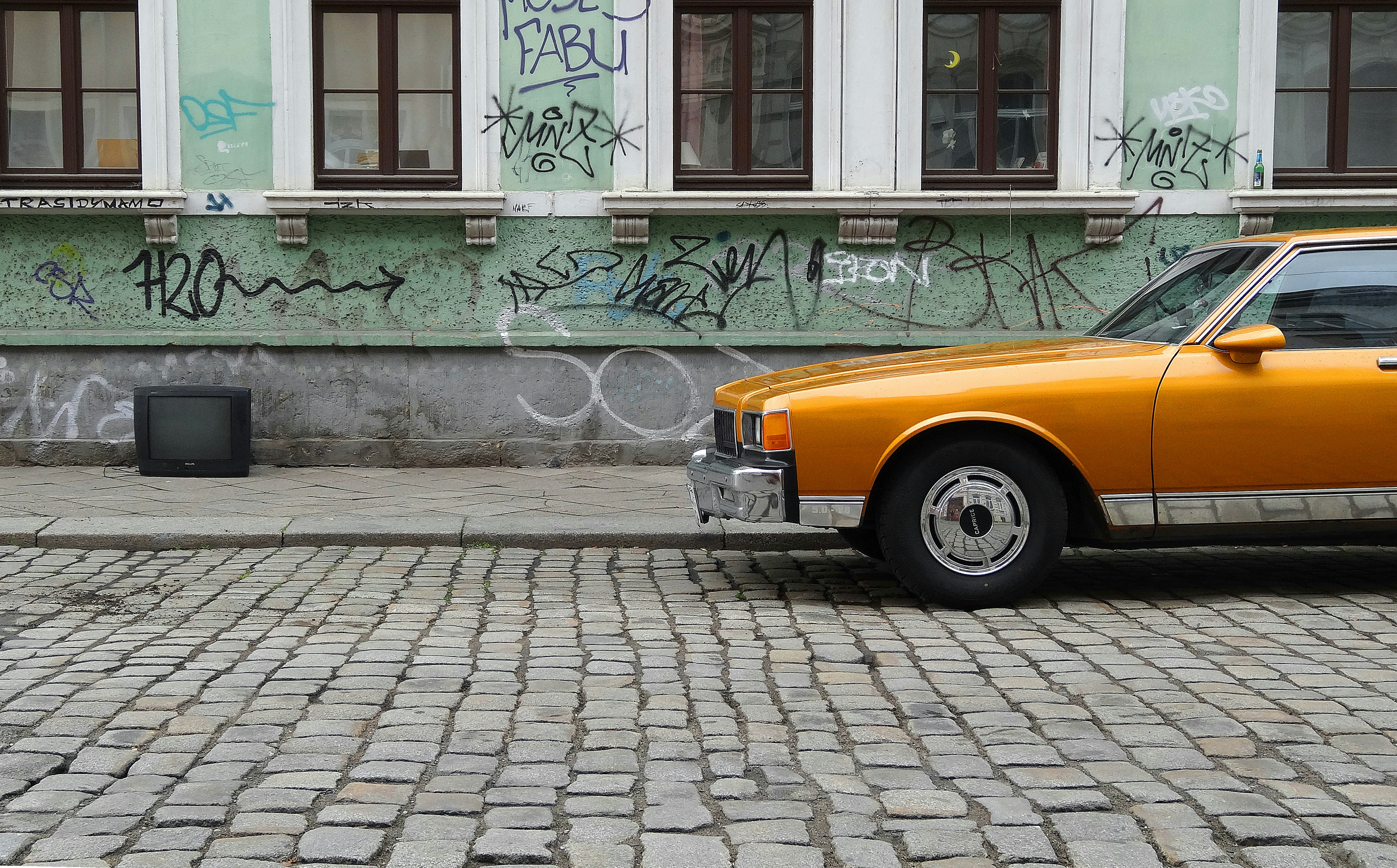 An orange vintage car parked beside a graffiti-covered wall, with an old television set on the cobblestone street.
