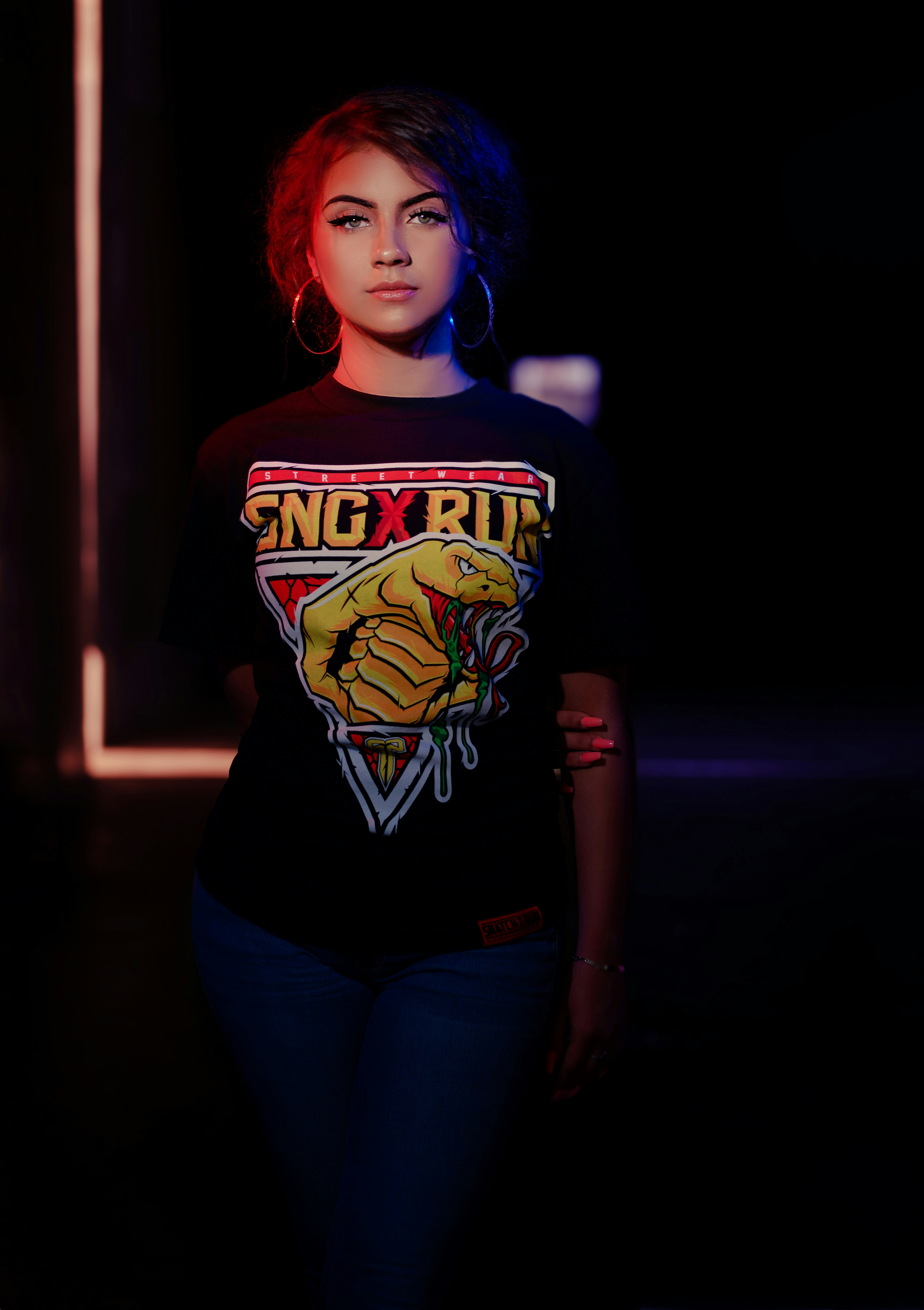 A young woman in a graphic t-shirt stands confidently in a dimly lit urban setting, illuminated by striking neon highlights.