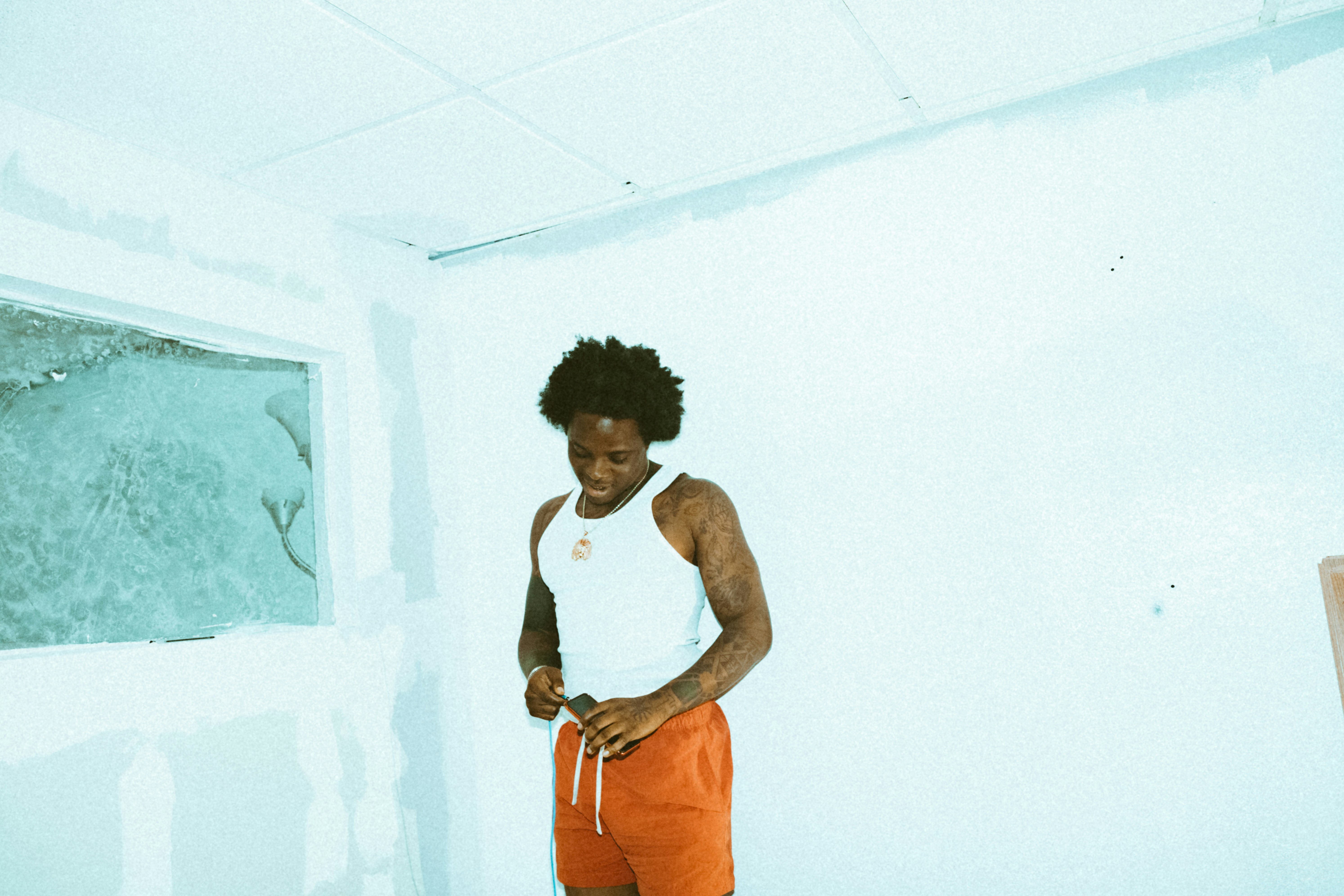 Man in white tank top and orange shorts stands in a bare, cyan-tinted room with a window to the left.