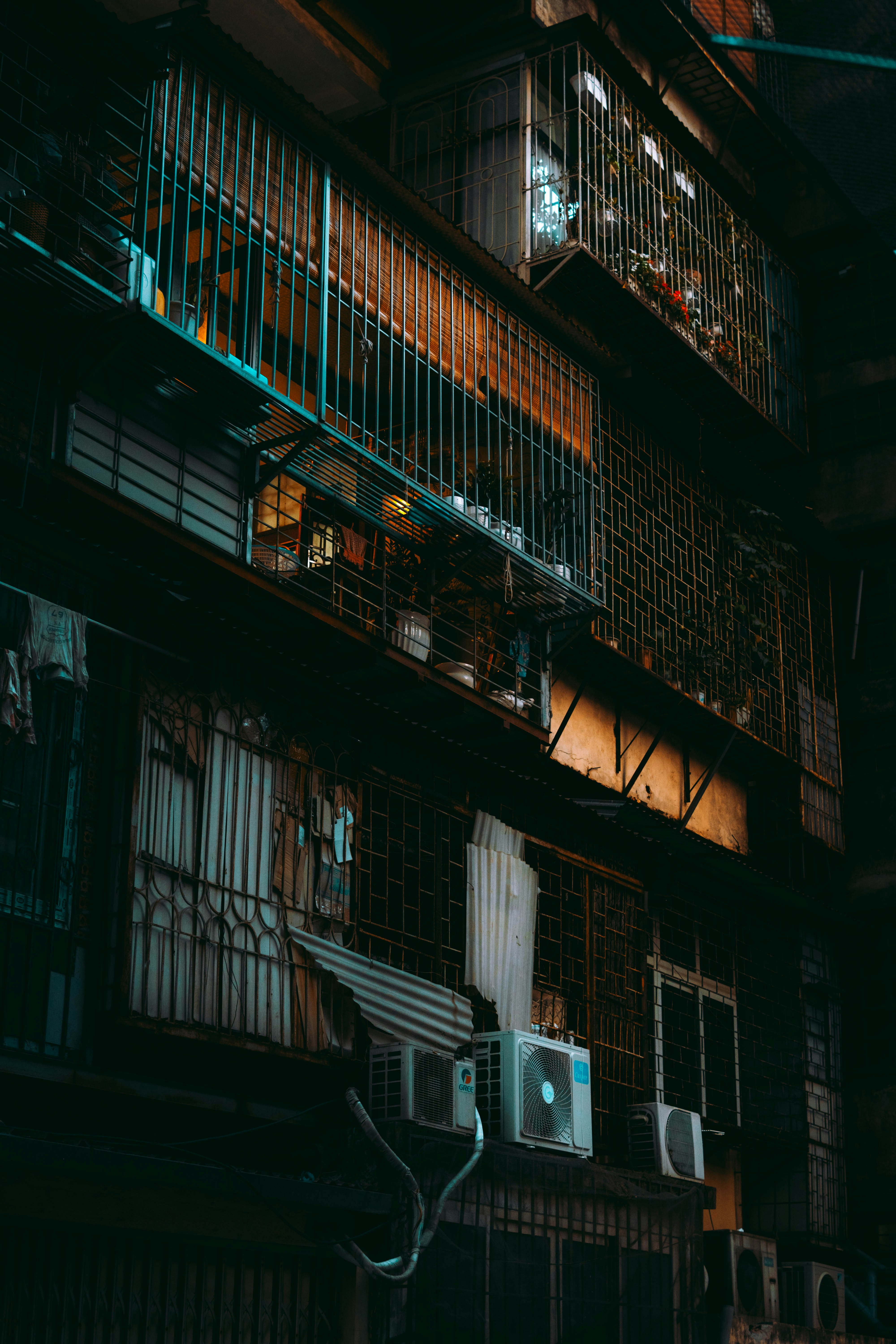 Dilapidated building facade showcasing weathered balconies and air conditioning units, illuminated by warm interior light.
