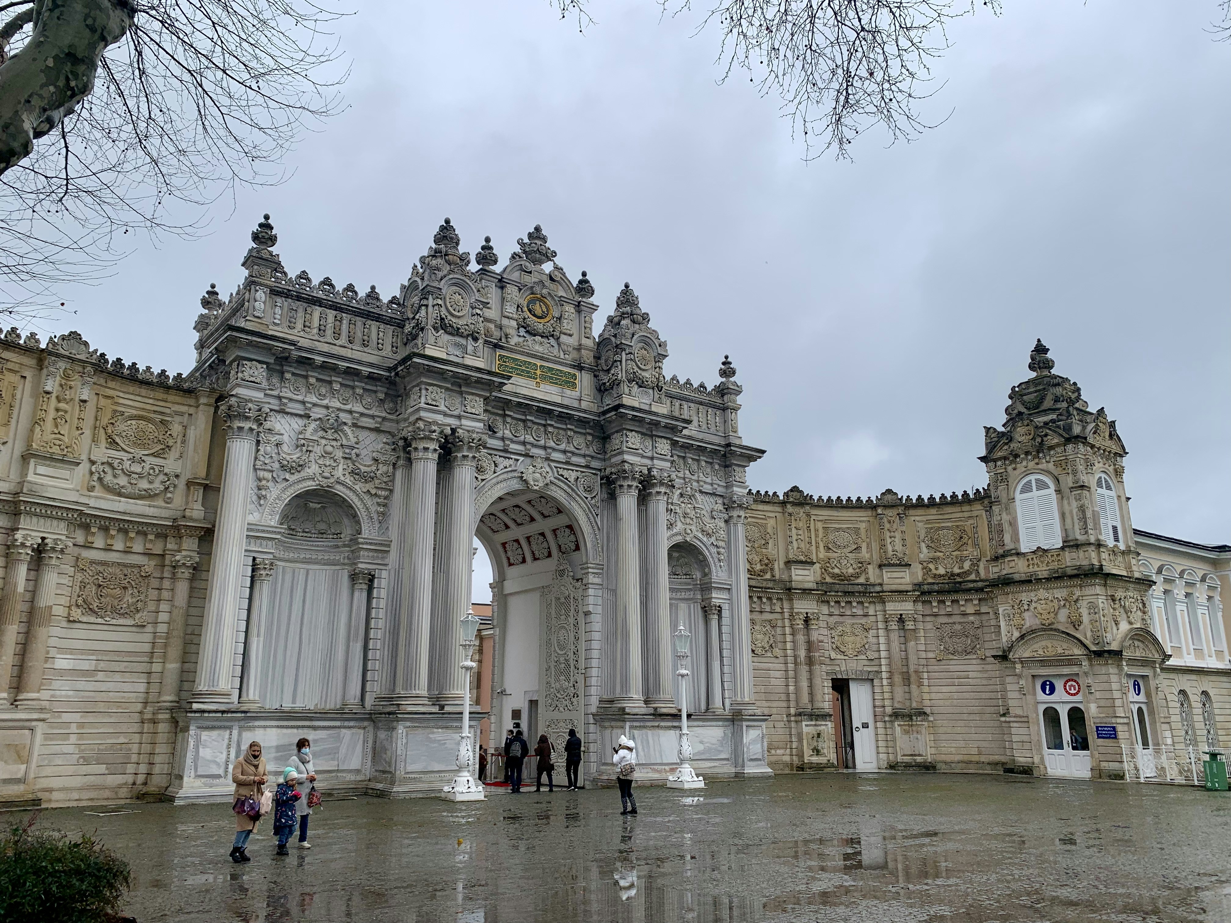 Elaborate architectural entrance of Dolmabahçe Palace, showcasing intricate details and grandeur. Visitors gather beneath the ornate archway amidst a rainy atmosphere.