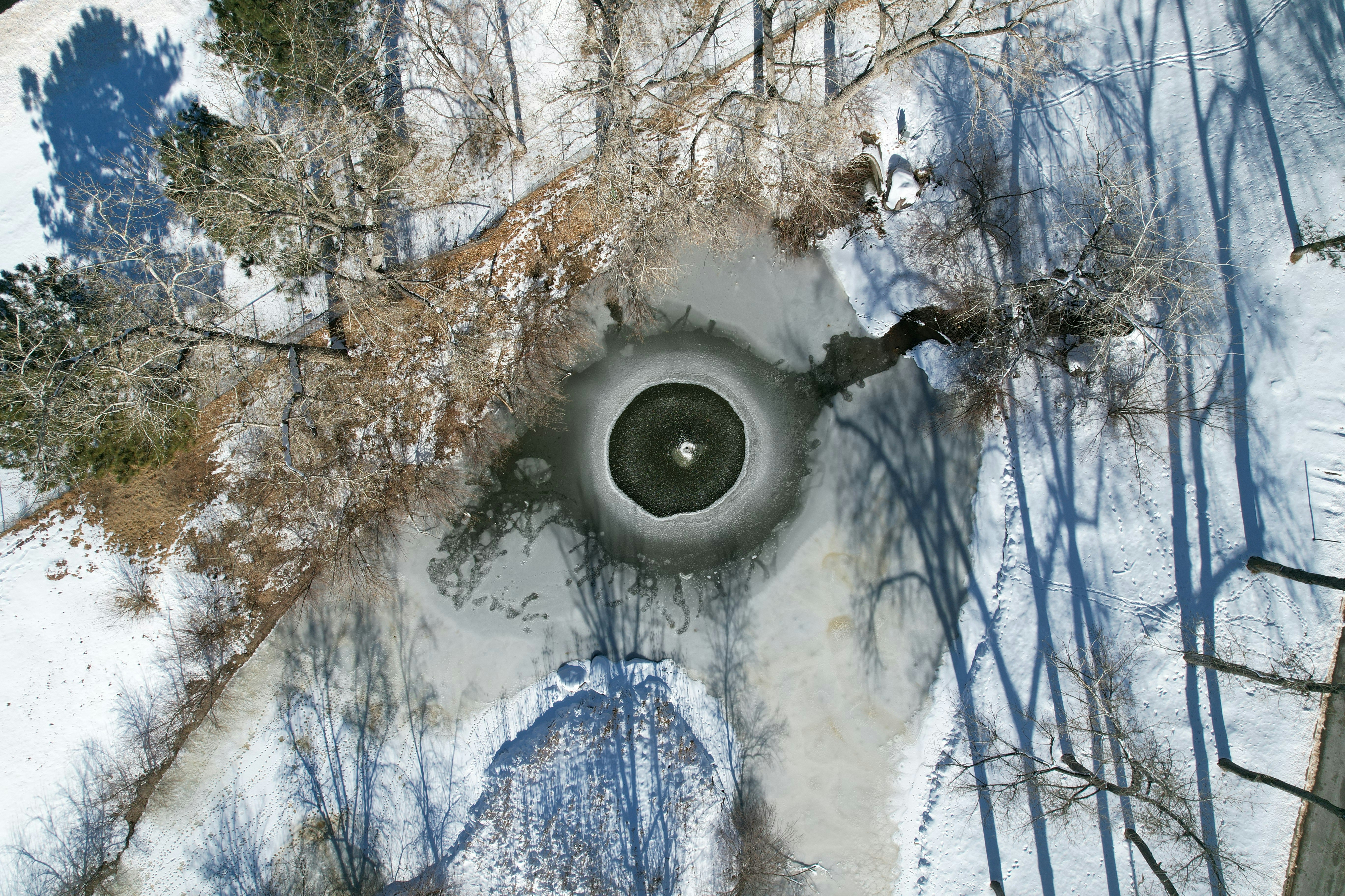 Aerial view of a circular area of open water surrounded by snow and ice, with long shadows cast by bare trees. The scene captures the contrast between the frozen landscape and the liquid core.